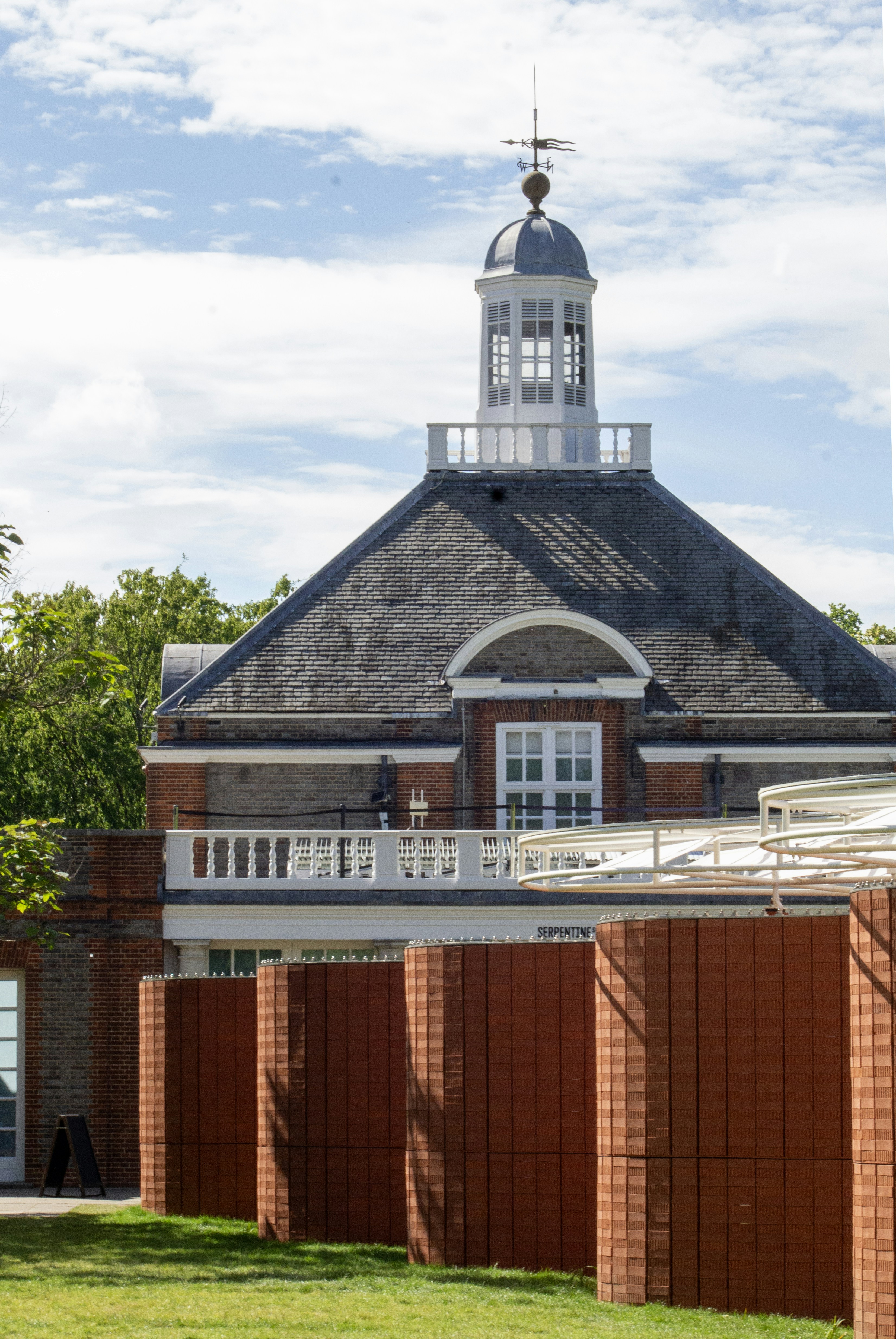Brick university-style building with a slate roof and cupola rises above a row of red brick columns; foreground shows a grassy lawn and a white railing.