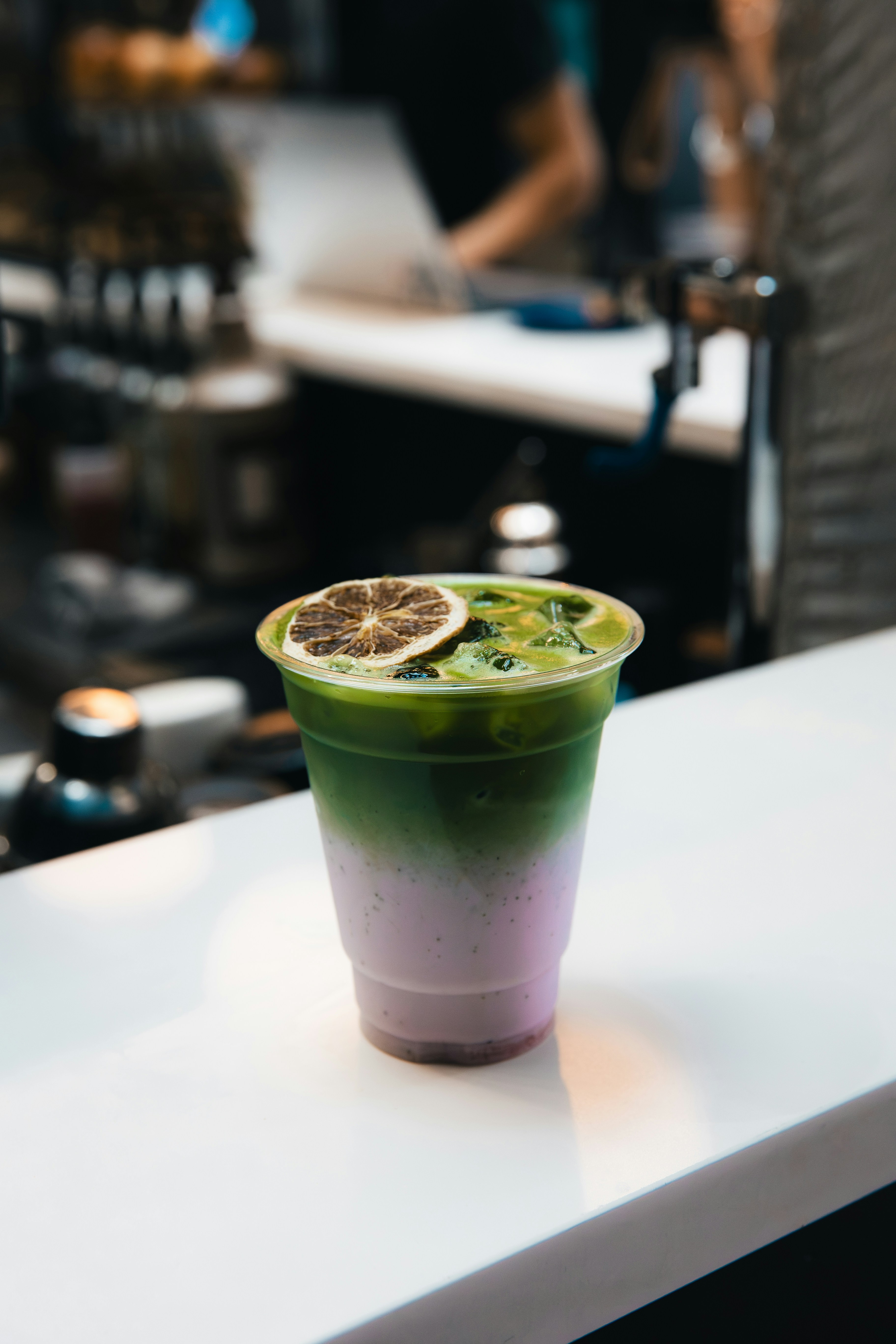 A vertical close-up captures an artisanal iced beverage featuring distinct layers of pastel lavender milk and vibrant green matcha. Topped with ice and a dried citrus wheel garnish, the drink rests on a sleek white cafe bar with a softly blurred espresso counter in the background.