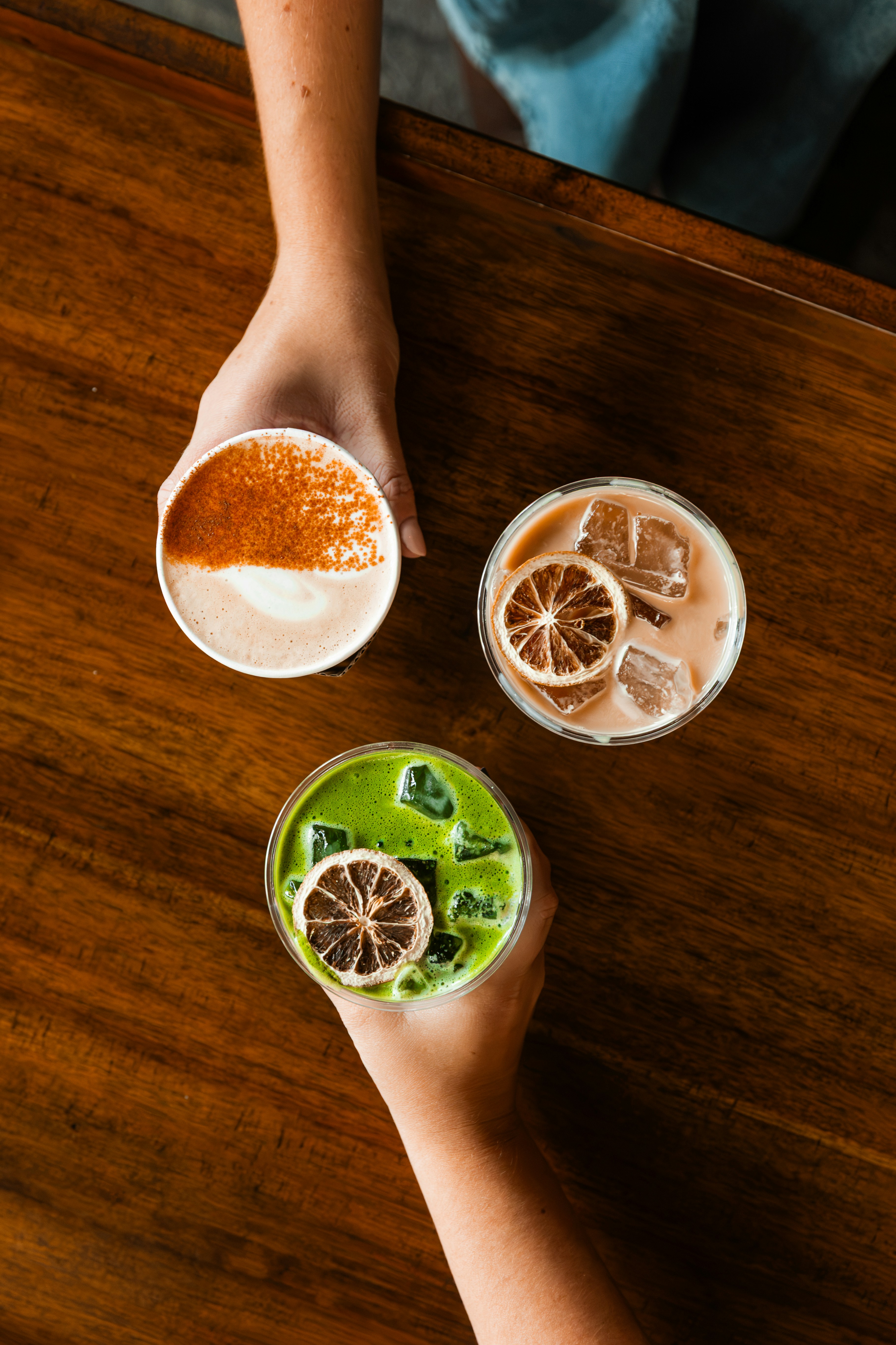 A top-down vertical perspective captures hands reaching for a vibrant trio of specialty cafe beverages arranged on a warm wooden table. The selection features an iced matcha latte and an iced chai both garnished with dried citrus wheels, alongside a frothy hot latte dusted with sweet cinnamon spice.