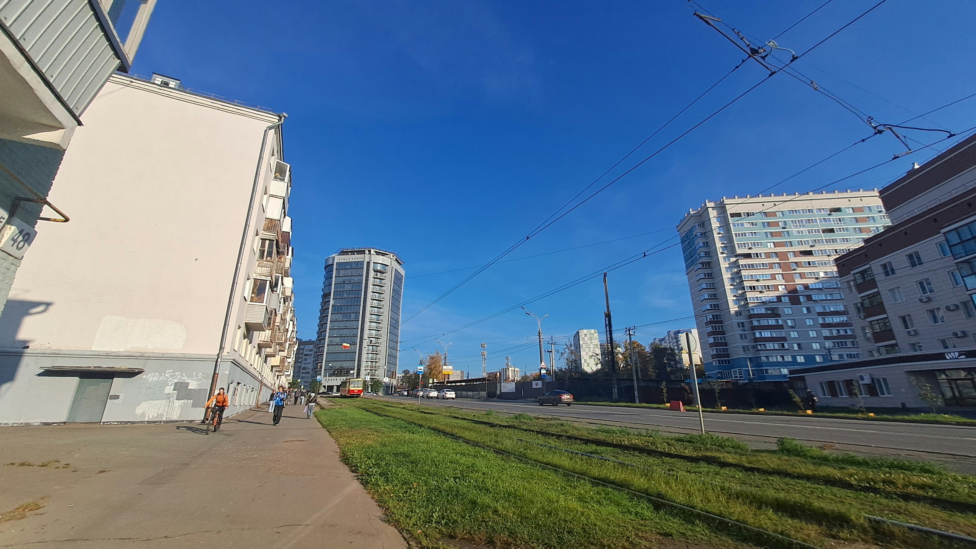 Wide street scene with modern apartment blocks and a clear blue sky; grass median and sidewalk hint at urban life and quiet morning activity.