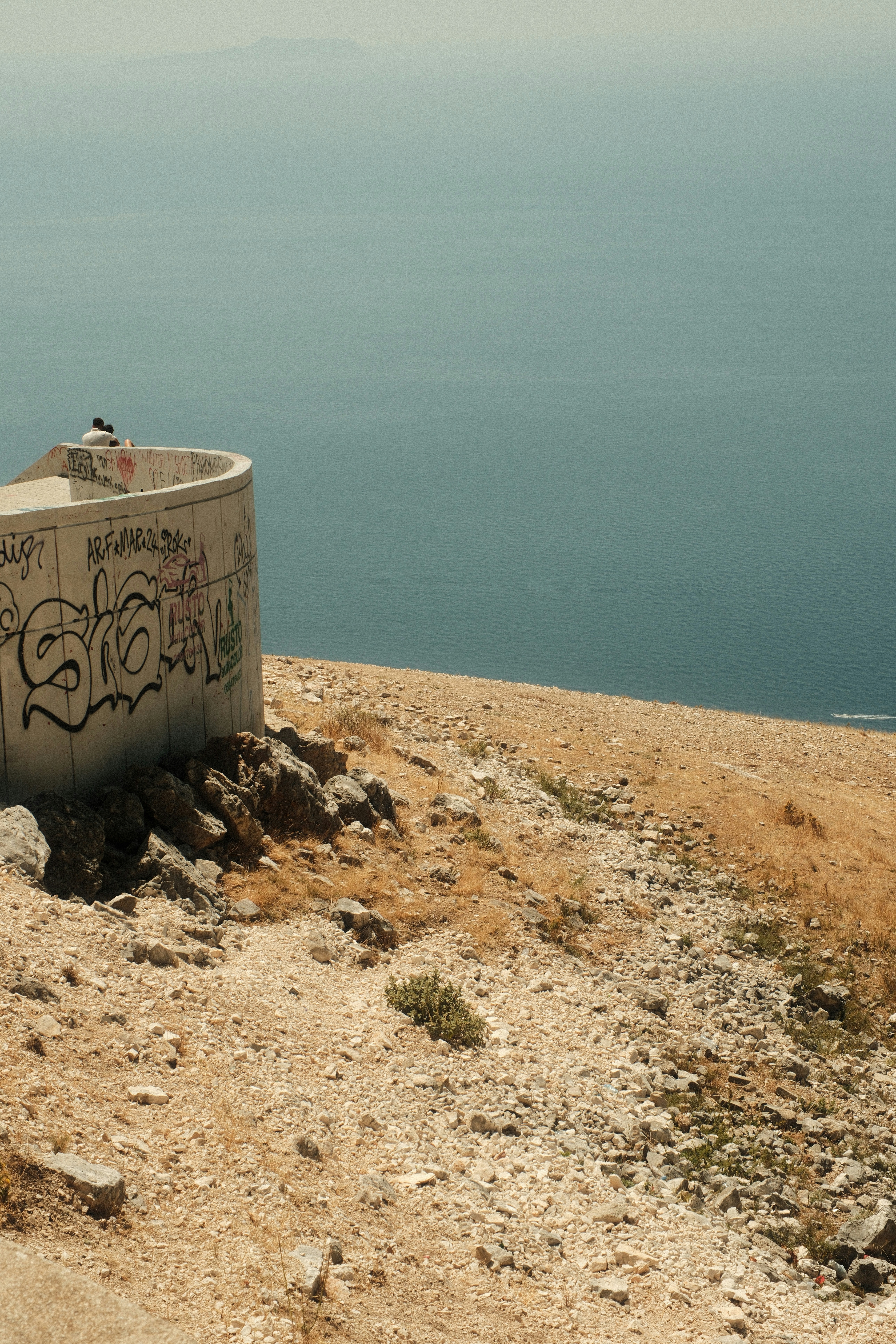 Two people on a graffiti-covered concrete wall overlooking the calm blue sea