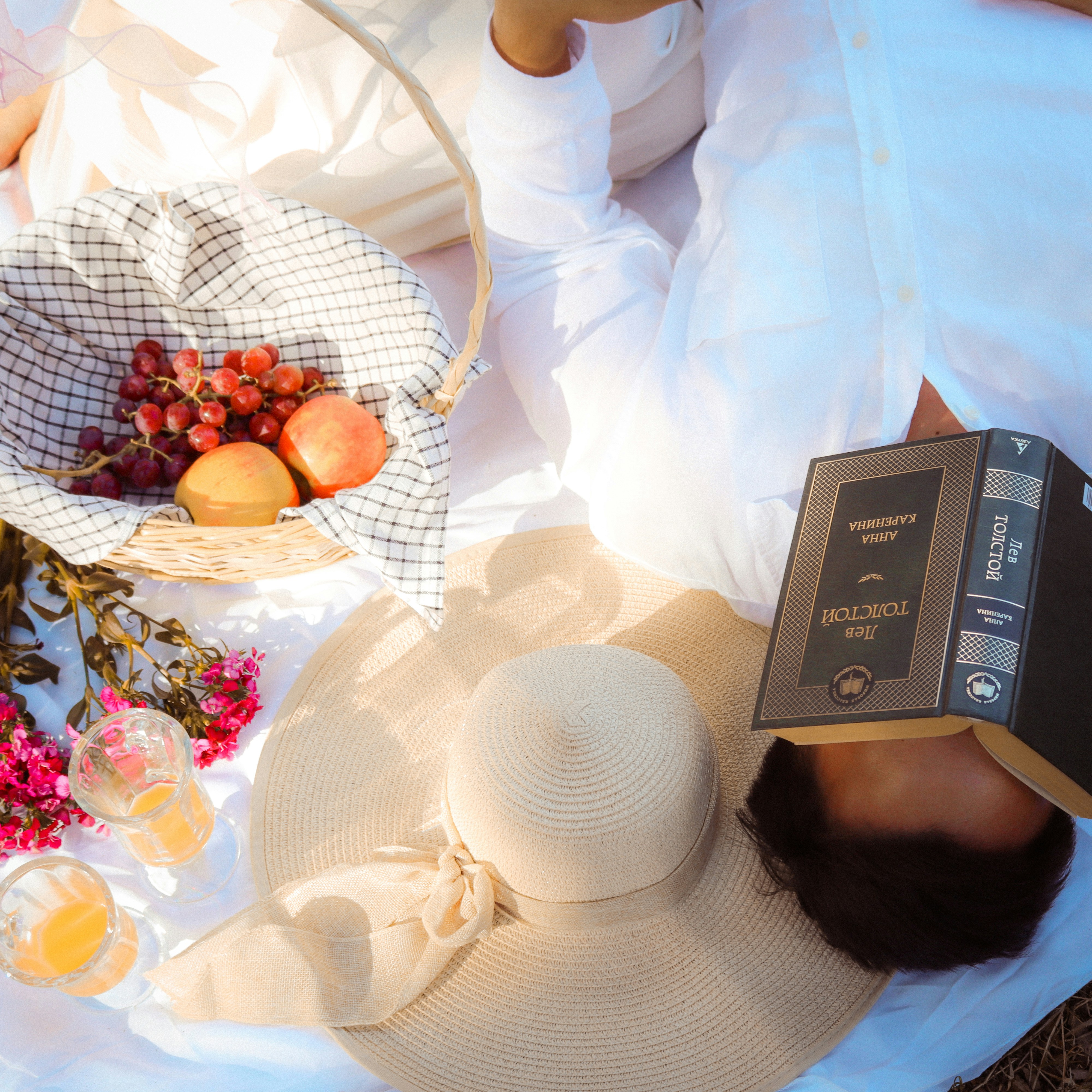 A relaxed scene of a person in white with a straw hat, a basket of fruit, and a book resting on the chest, captured during a sunlit picnic. The image shows casual detail and warm tones.