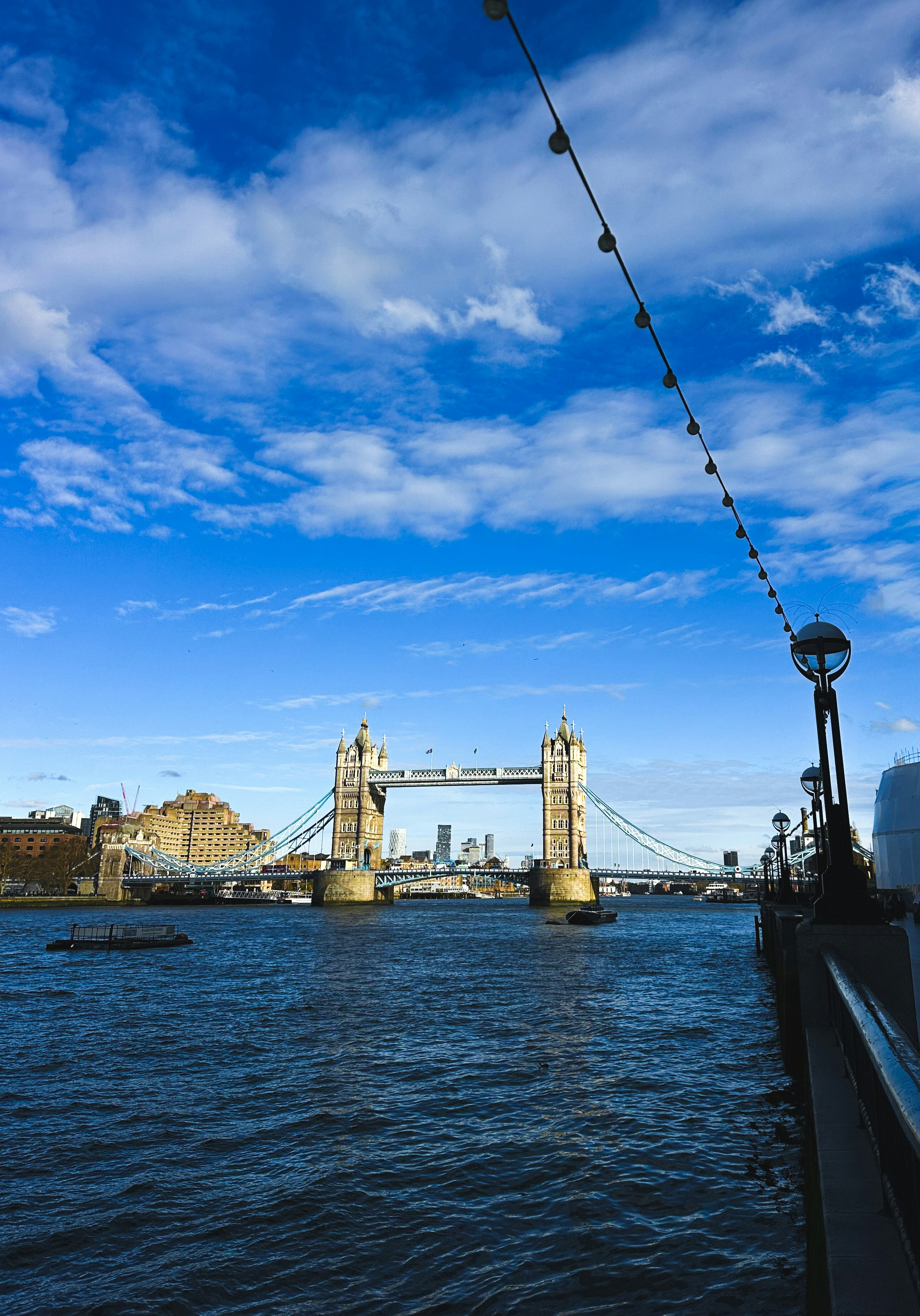 A striking view of Tower Bridge spanning the River Thames in London, framed by dramatic clouds and riverside architecture. The deep blue sky, flowing water and distinctive riverside street lights create a classic London cityscape.