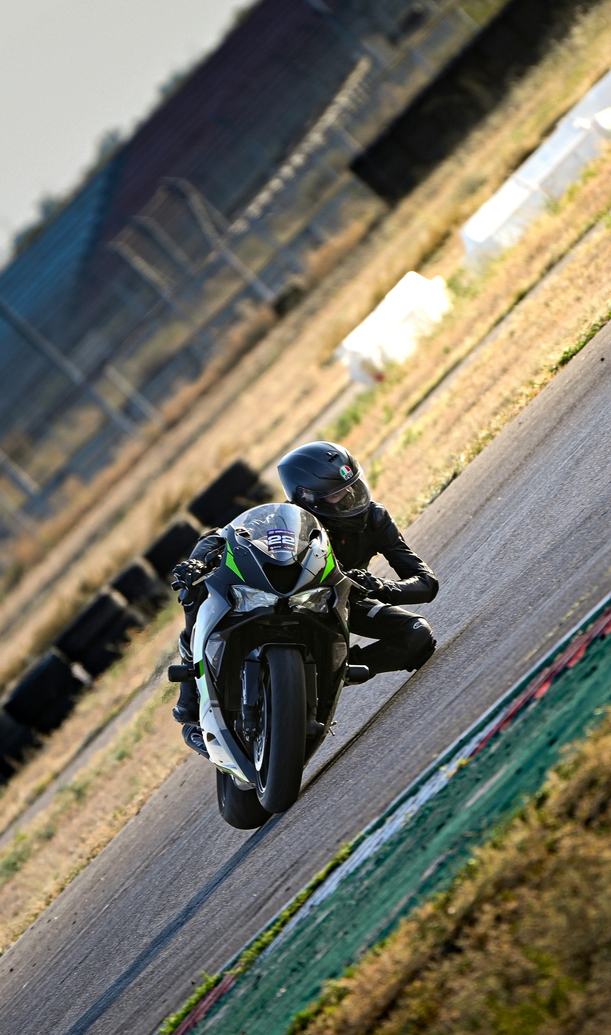 Rider on a sport bike leans through a corner on a paved track, wearing a black helmet and suit with a blurred industrial backdrop.
