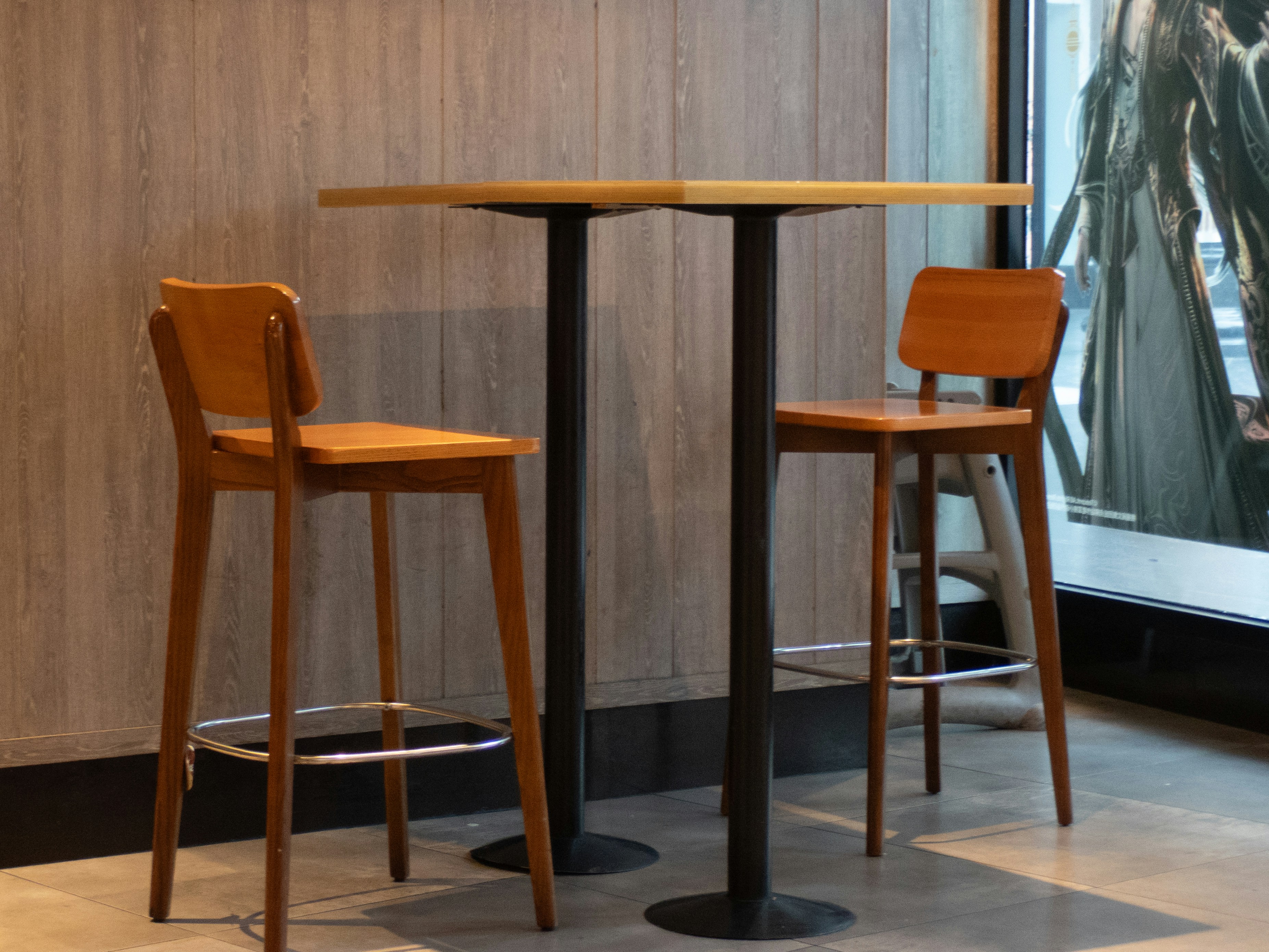 Two wooden stools flank a tall table in a quiet café, with a wooden-paneled wall and large glass window in the background.