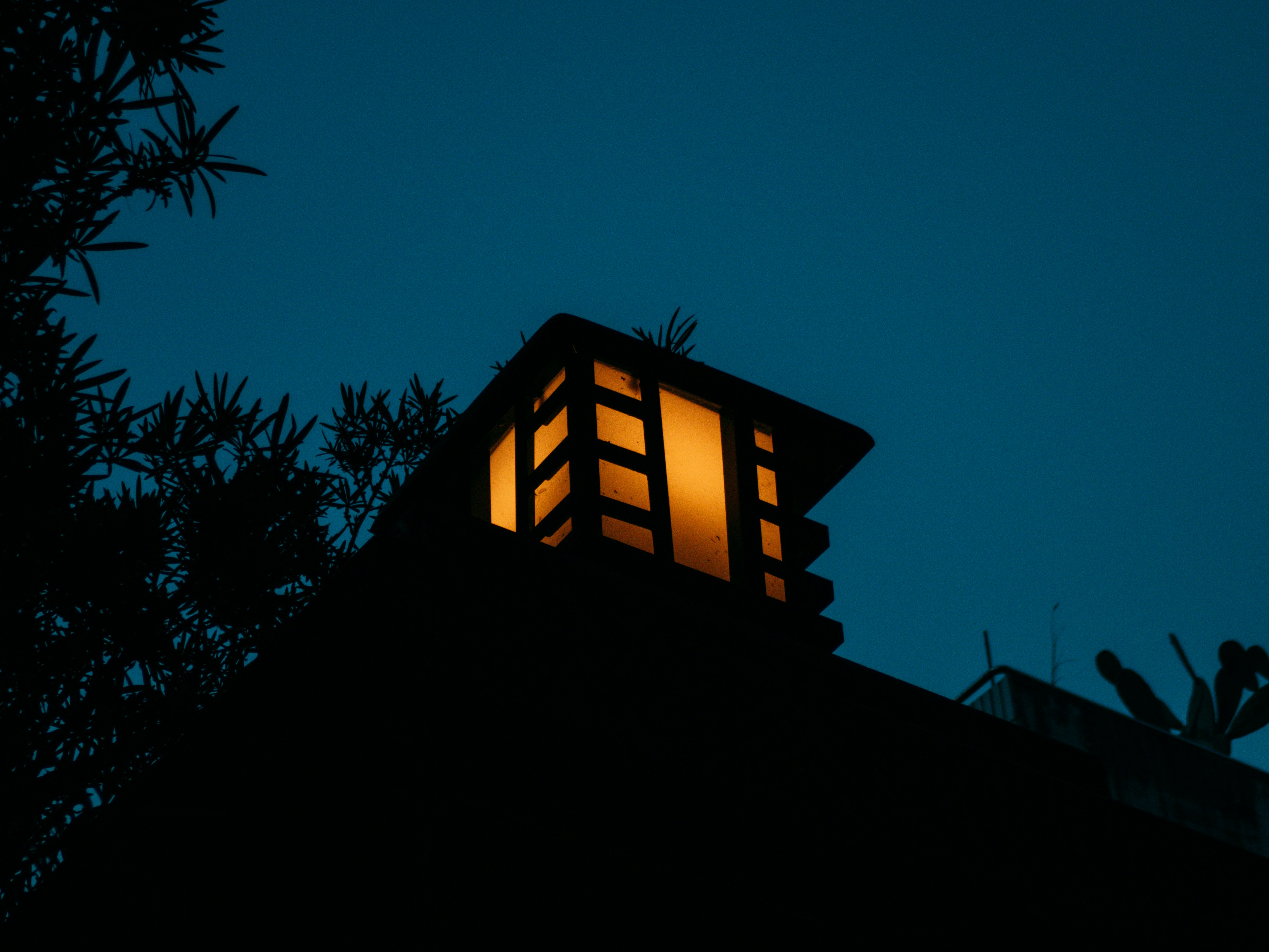 Dimly lit lantern glows from a roof crest against a night sky, framed by silhouettes of foliage and building edges. An architectural vented lantern emphasizes warm light in a moody urban rooftop scene.