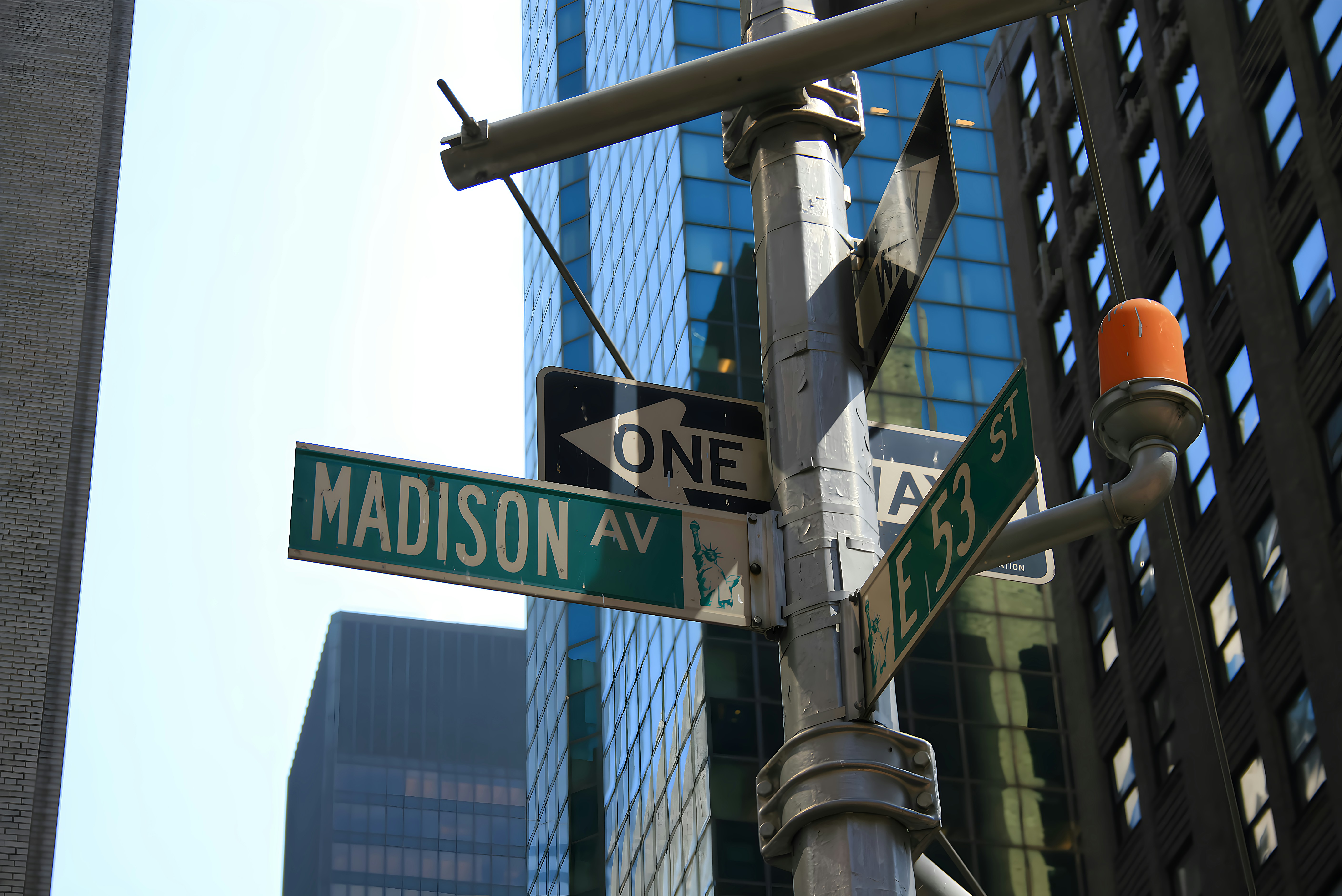 Street corner signs for Madison Ave and East 53rd St against modern glass towers; urban intersection with a traffic signal in view.
