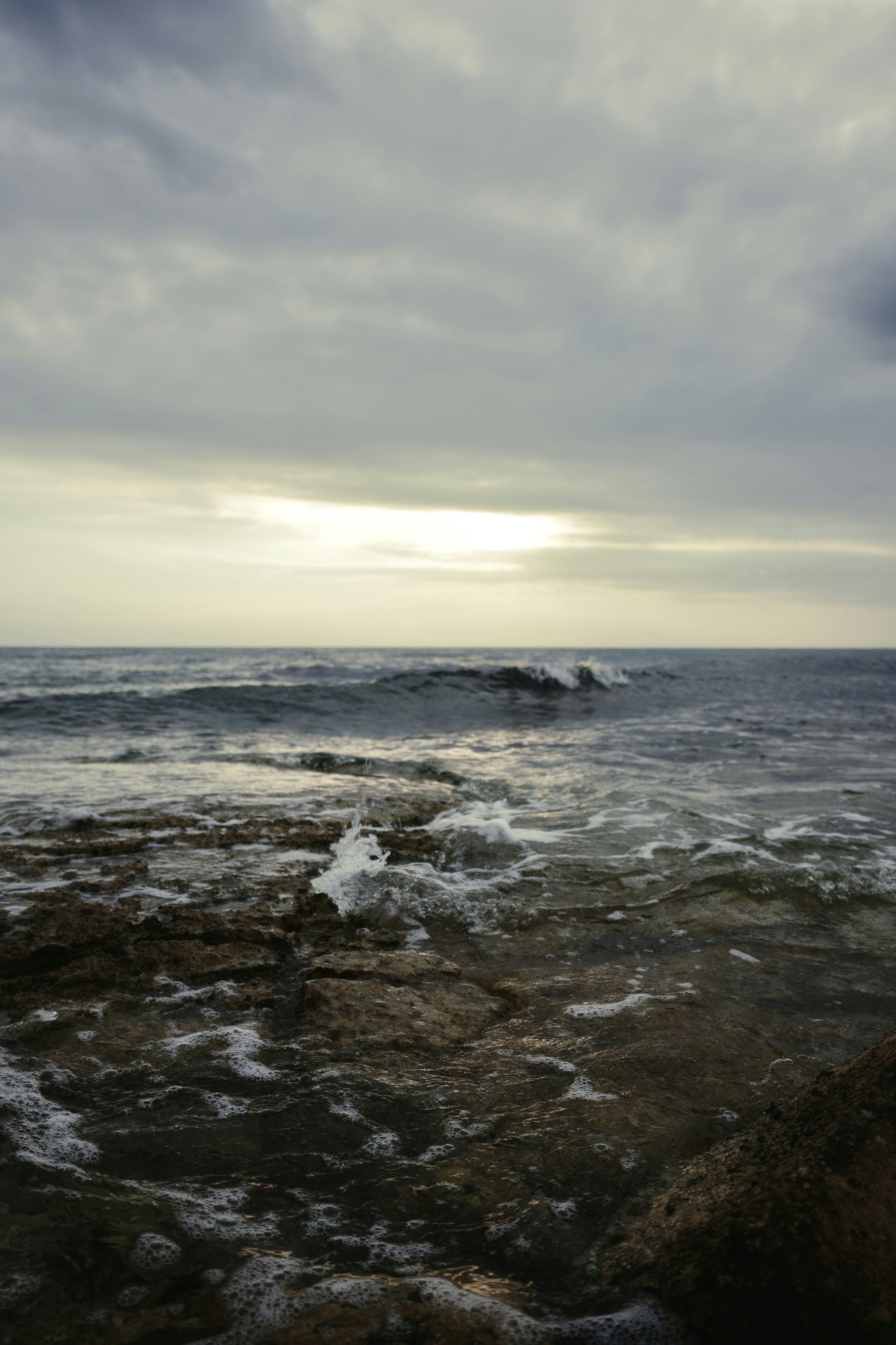 Coastal rocks meet foaming waves beneath a cloudy sky, with a soft glow on the horizon. The scene emphasizes the sea’s texture and the rugged foreground.