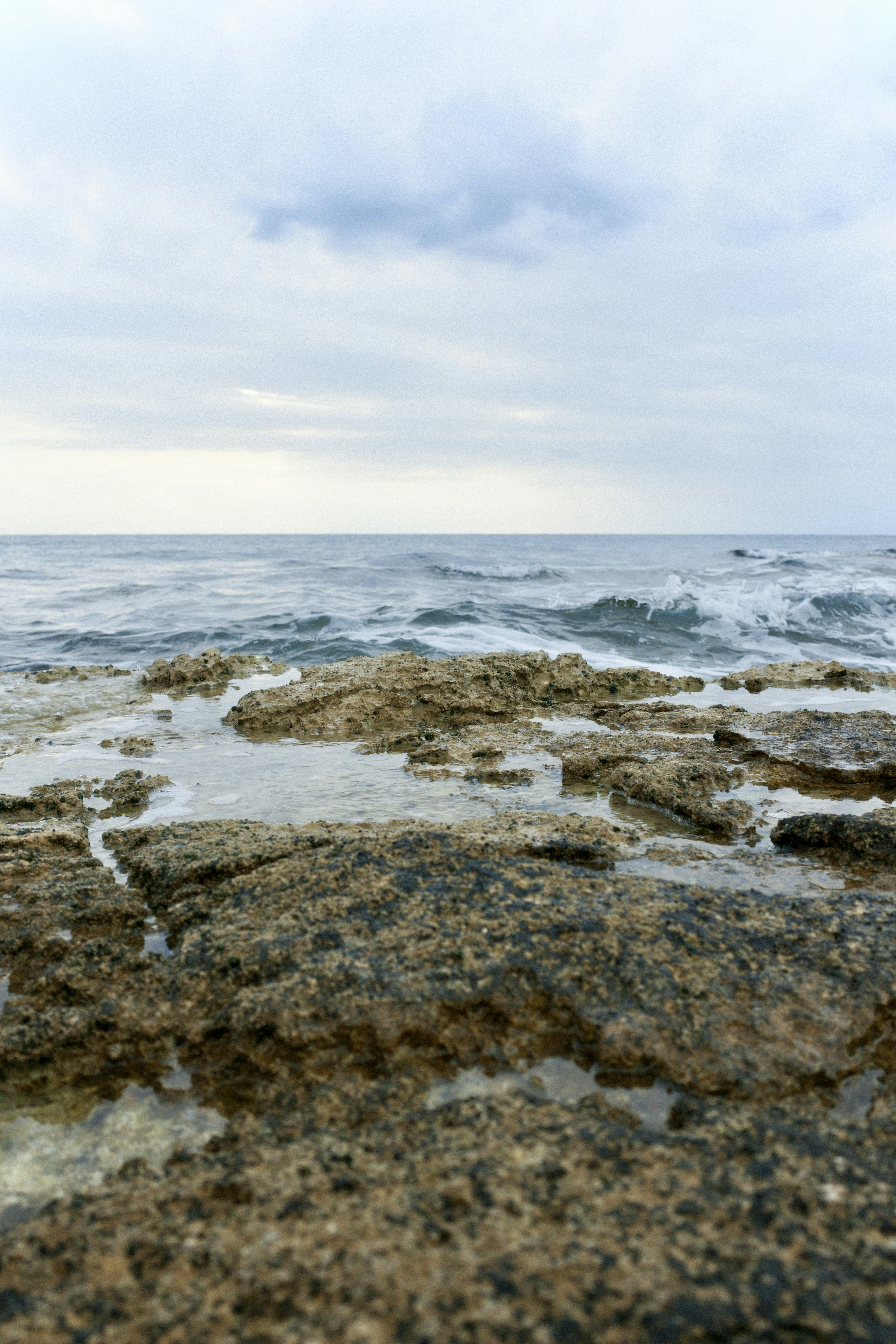 Rocky shoreline with waves approaching, distant horizon under overcast sky; a tranquil coastal scene with wet stones in the foreground. This is a landscape photograph, capturing texture and depth.