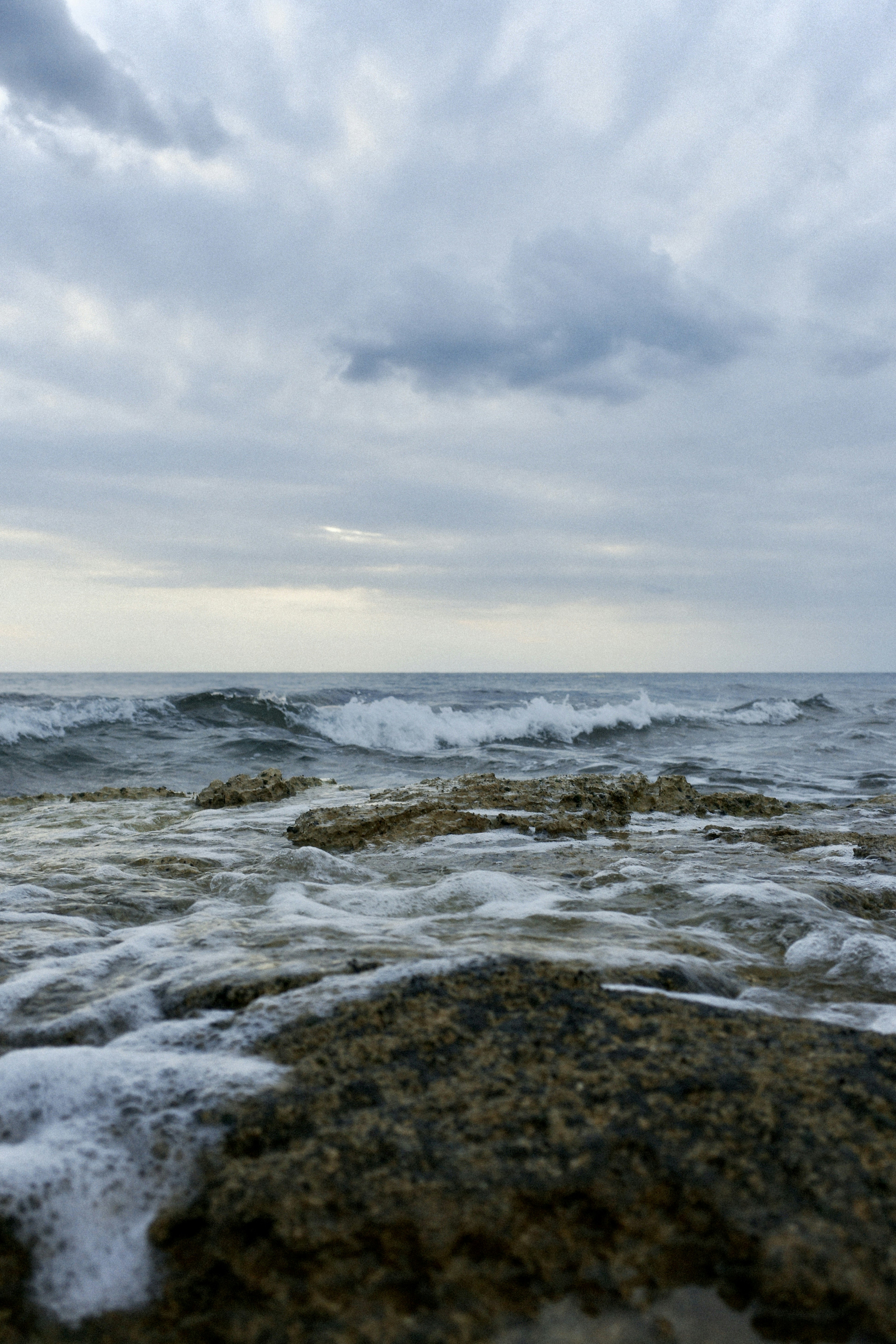 Rugged shoreline with foamy surf and textured rocks under a cloudy sky, capturing the sea’s motion and muted coastal tones.
