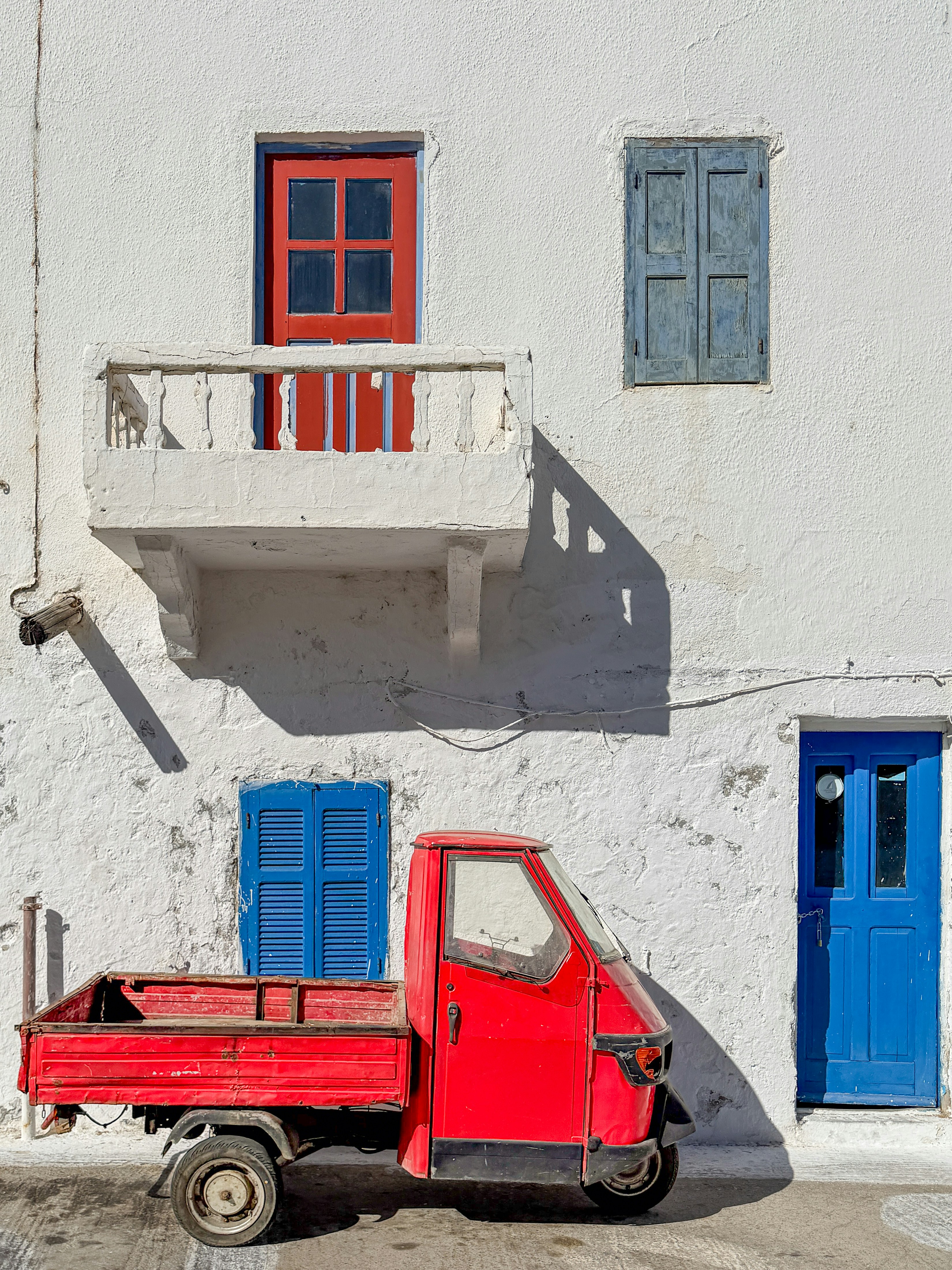Bright red utility tricycle sits against a sunlit white wall with blue doors and a red-framed window, capturing a Mediterranean-town street scene.
