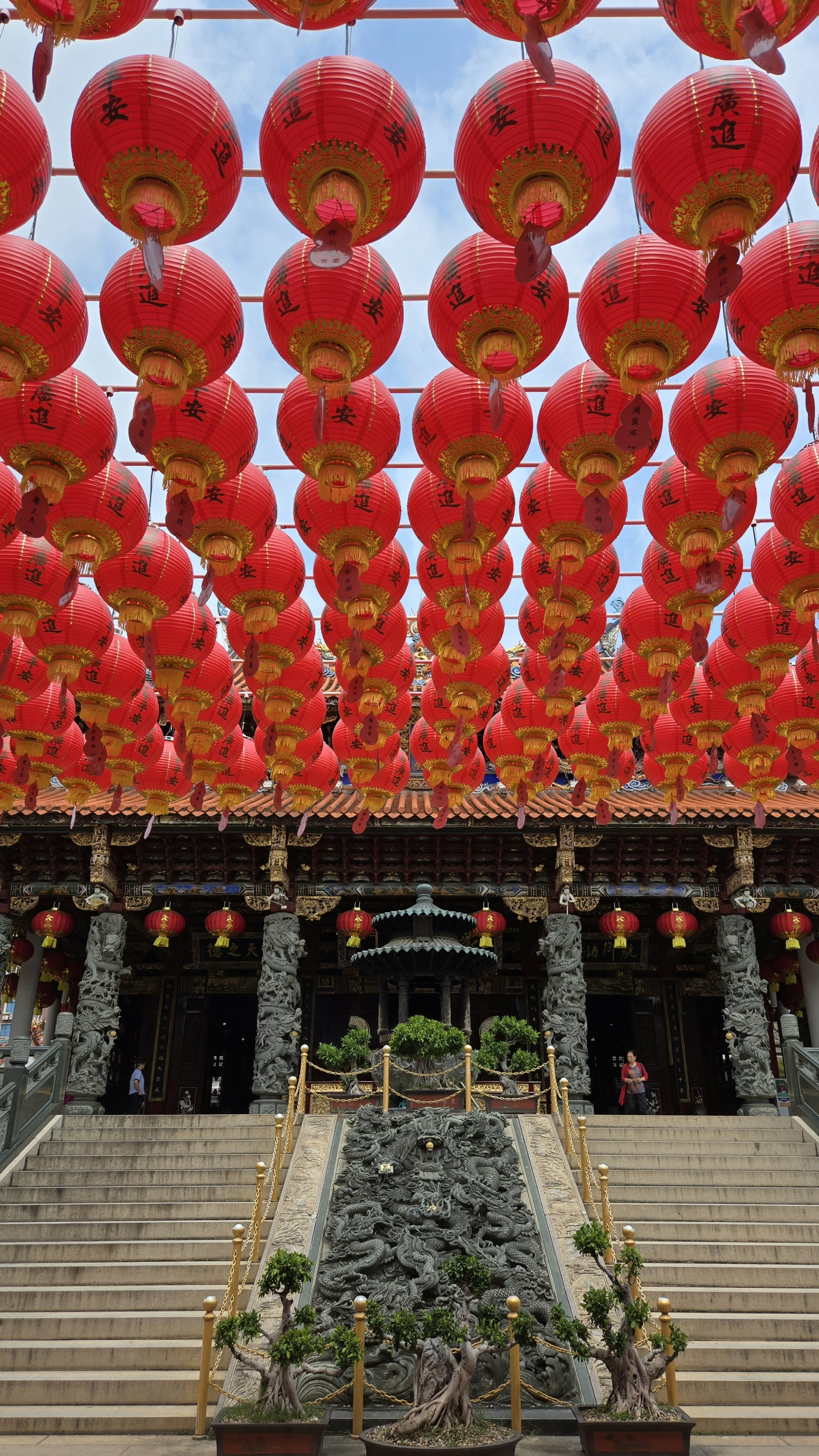 Red lanterns hang in neat rows above a temple courtyard with carved stone railings and steps leading to an ornate entrance.
