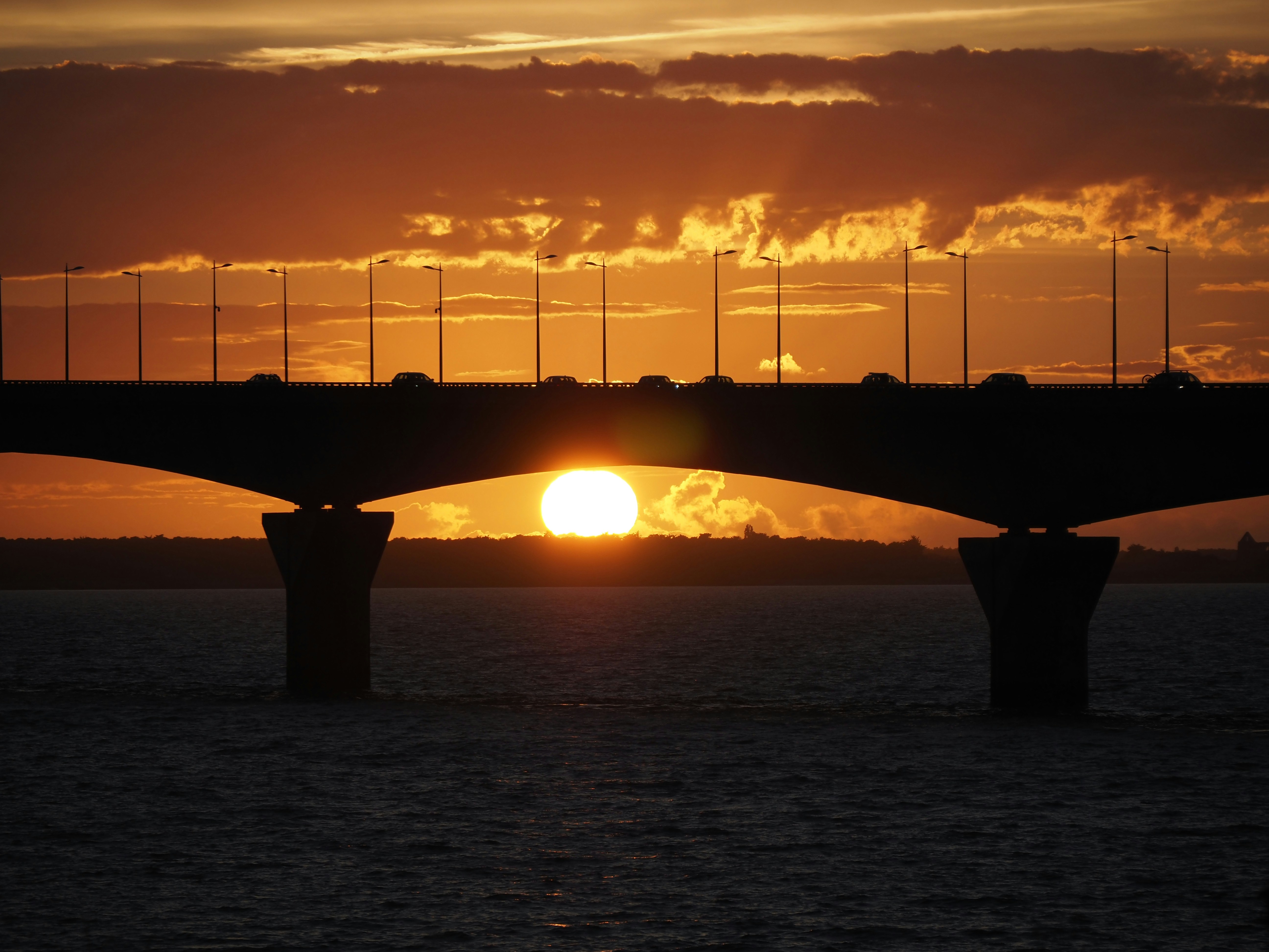 Golden sunset settles behind a silhouetted bridge, casting a warm glow on the water below. A calm, horizon-to-horizon scene capturing the bridge’s silhouette and the fiery sky.