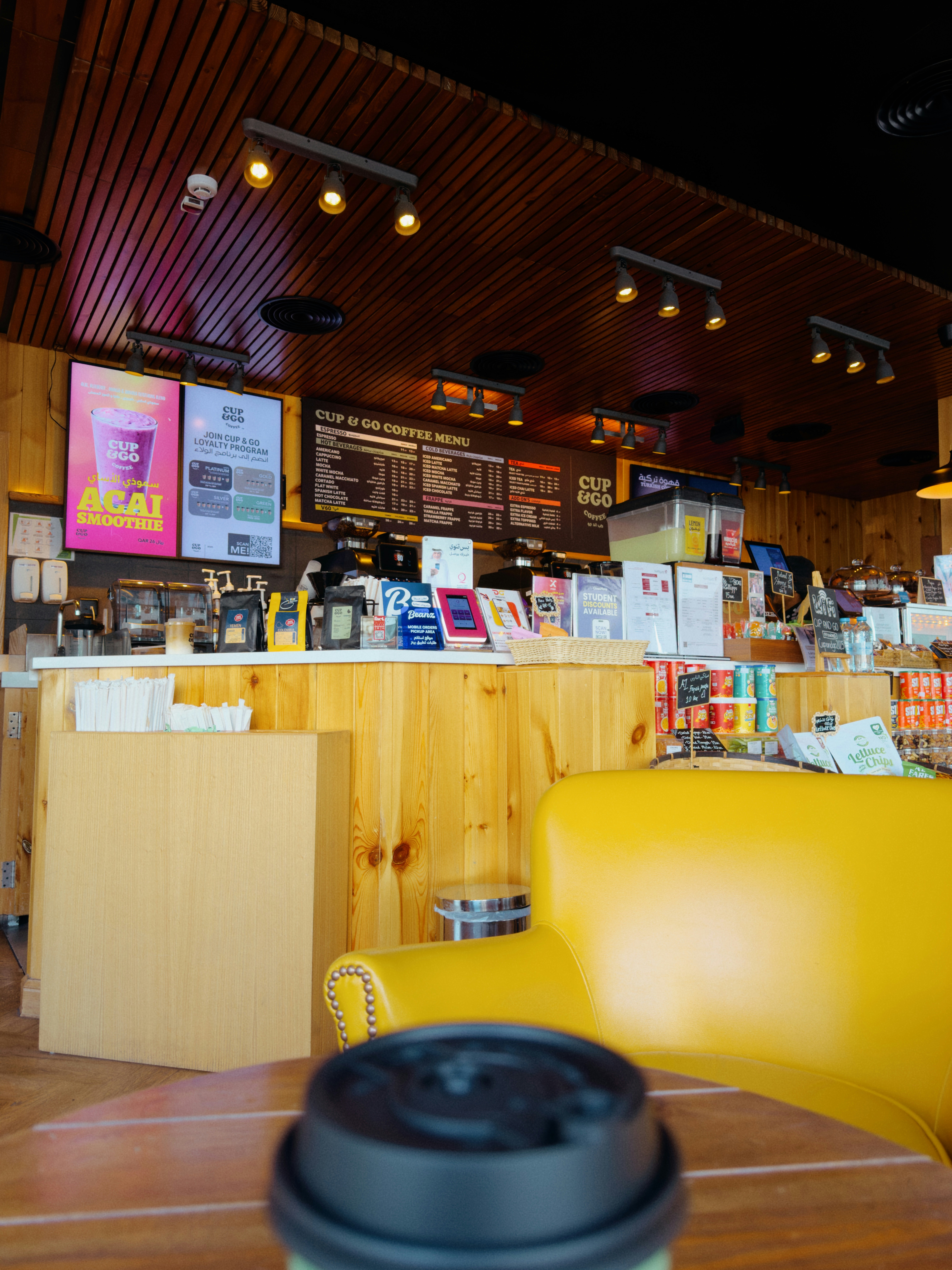 Bright interior of a cafe with a wooden counter and yellow leather seating; foreground shows a coffee cup lid on a table, while the back wall features menu boards and takeaway displays.