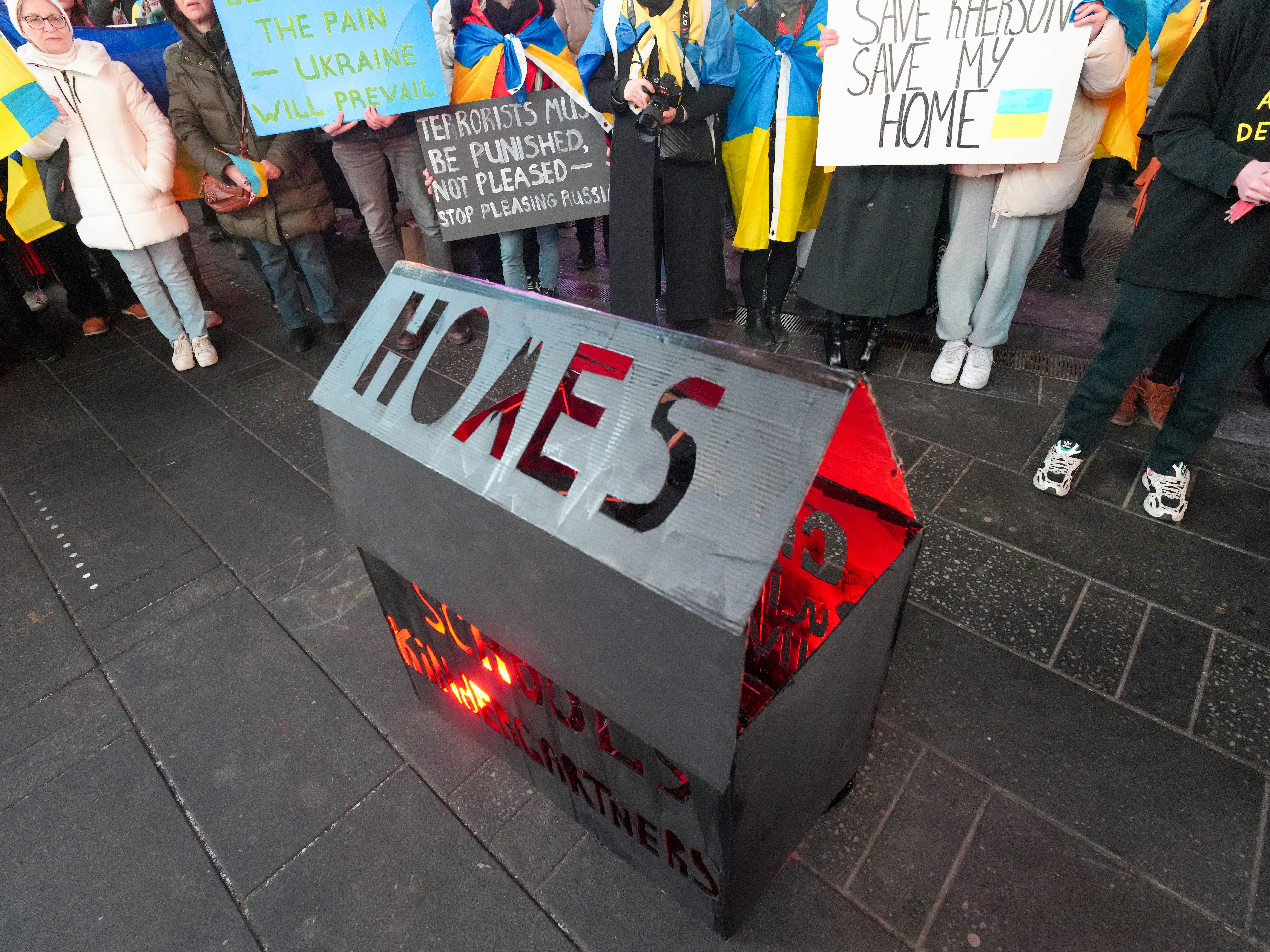 A black cardboard house sculpture with the word "HOMES" cut from its roof glows red from inside, set on the pavement in front of protesters. Demonstrators wrapped in Ukrainian flags hold signs like "Save Kherson, Save My Home" and "Ukraine Will Prevail." Protesters rally in Times Square, New York City, on February 15, 2022, standing in solidarity with Ukraine and speaking out against the war at an anti-war demonstration. Photo: Olek Buzunov (@buzunov.photoart)