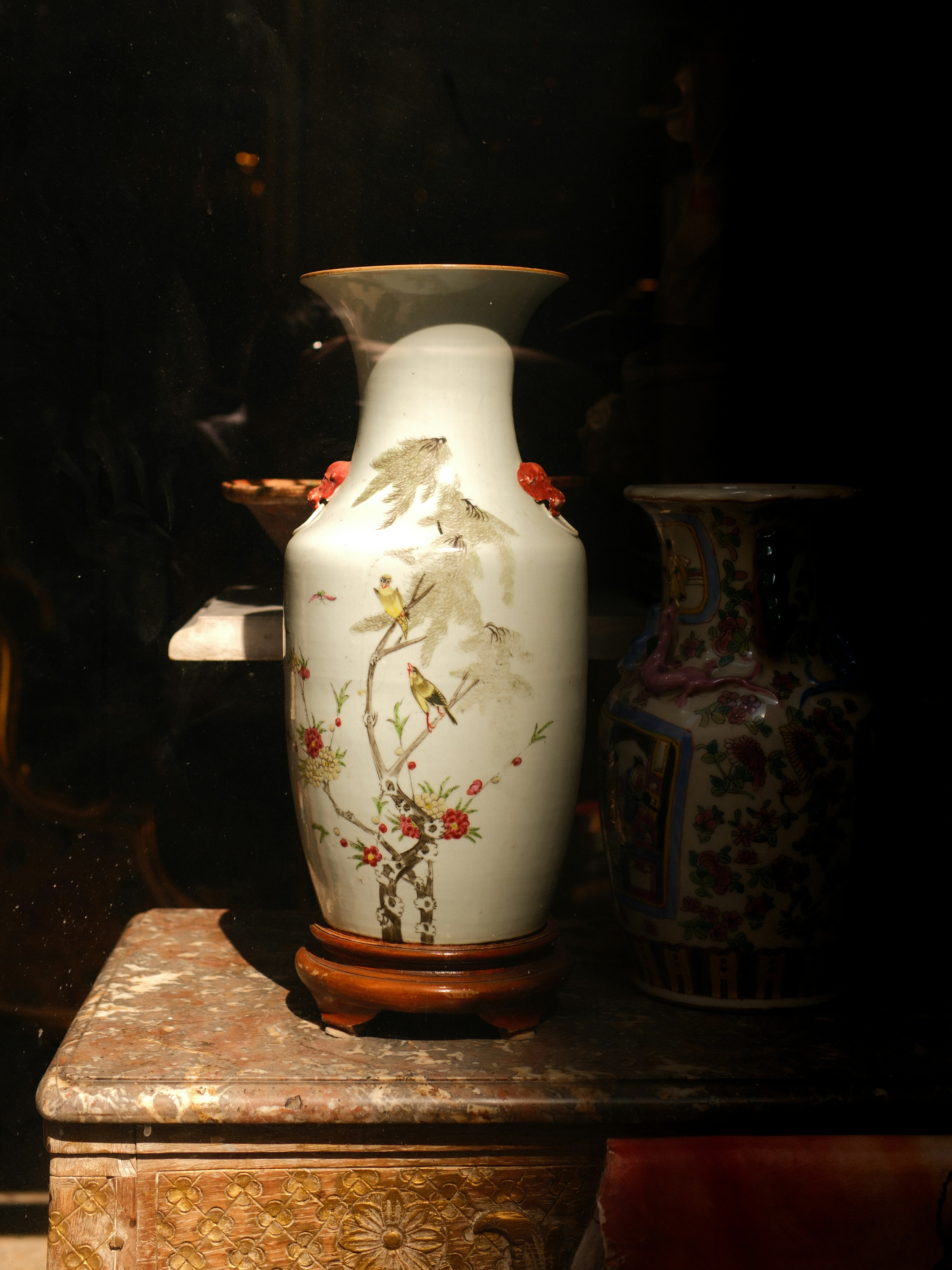 Porcelain vase with floral and bird motif placed on a carved wooden stand, illuminated against a dark backdrop in a museum-like display.