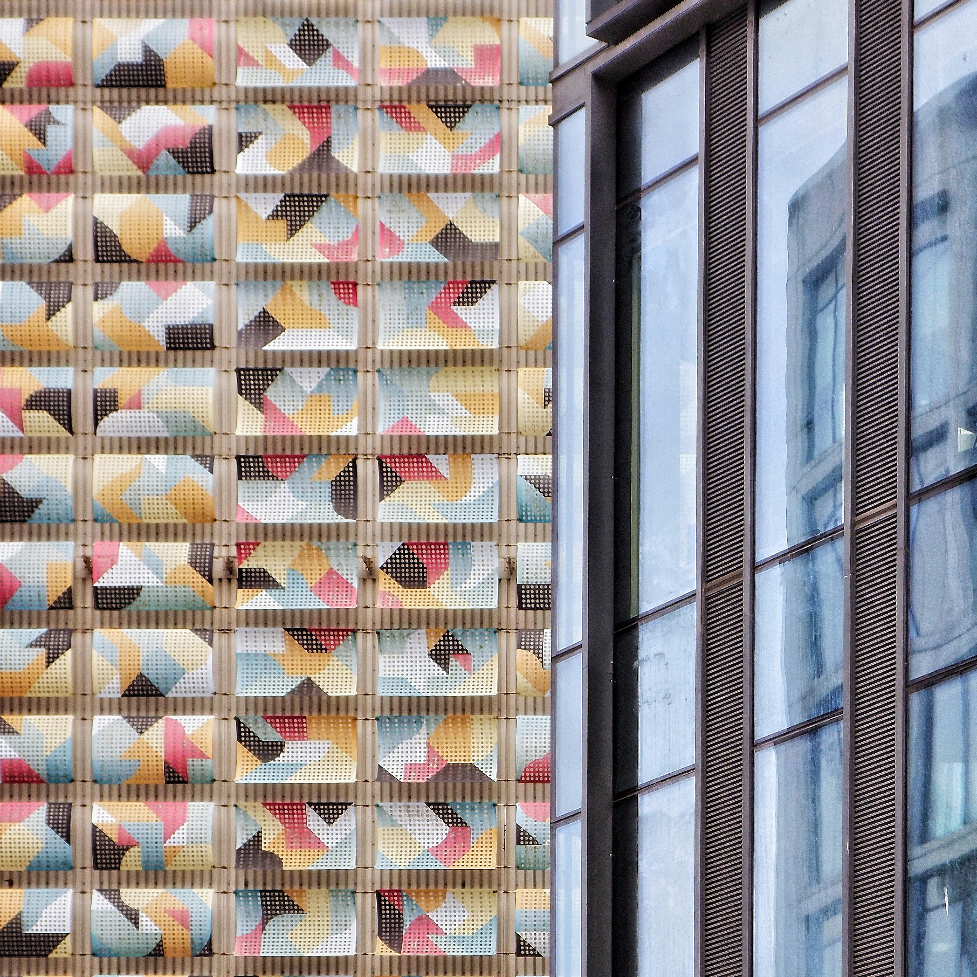 Colorful geometric panels decorate a building façade beside a tall glass windowed wall, juxtaposing tactile pattern with sleek urban glass.