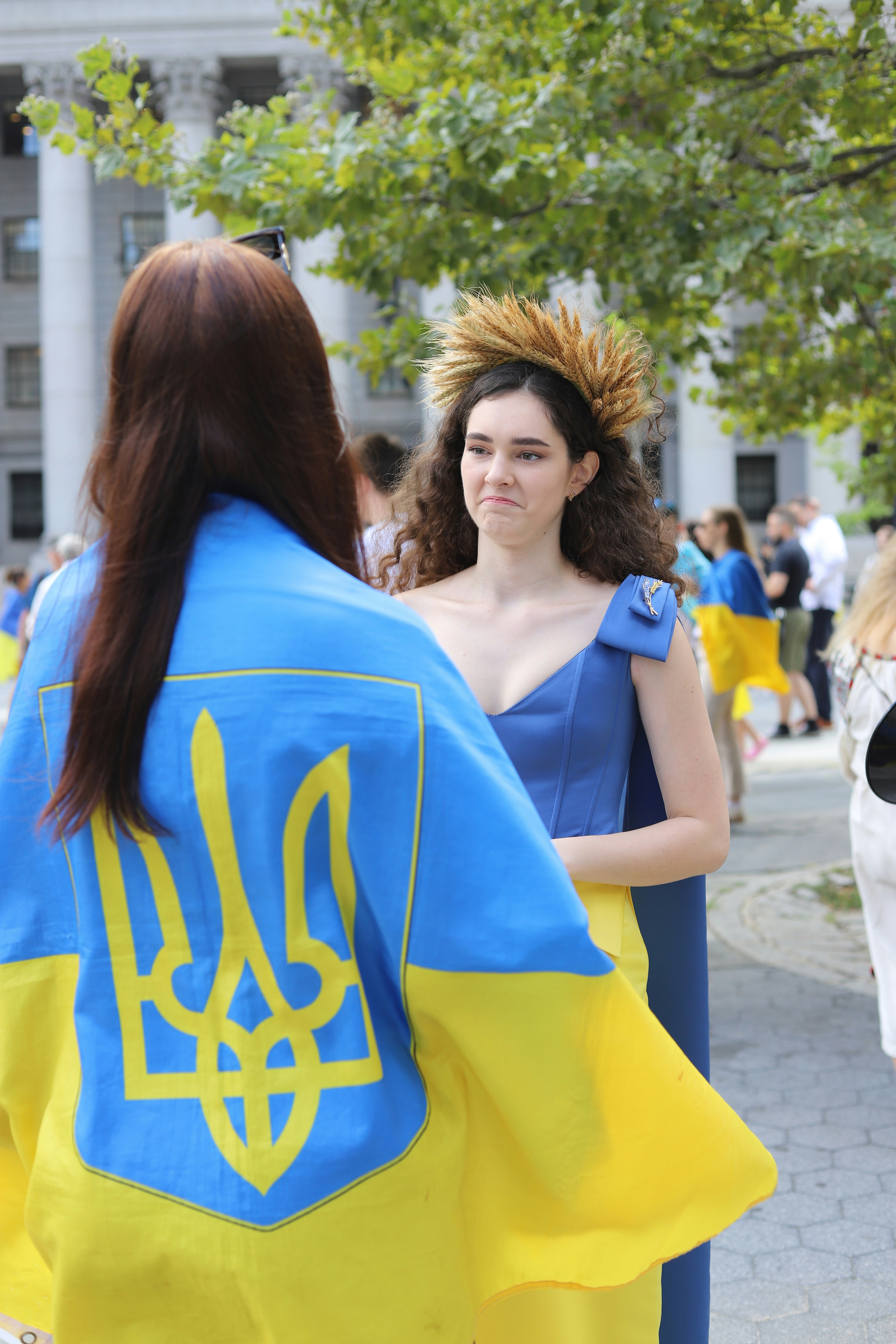 A young woman with curly dark hair wears a golden wheat crown and a blue off-shoulder dress at a Ukrainian rally in downtown Manhattan. Beside her, another woman is draped in a large blue and yellow Ukrainian flag with the trident emblem, with trees and buildings behind them. People gather in downtown Manhattan for a Ukrainian rally, waving blue and yellow flags and showing solidarity and support for Ukraine in the streets of New York City. Photo: Olek Buzunov (@buzunov.photoart)