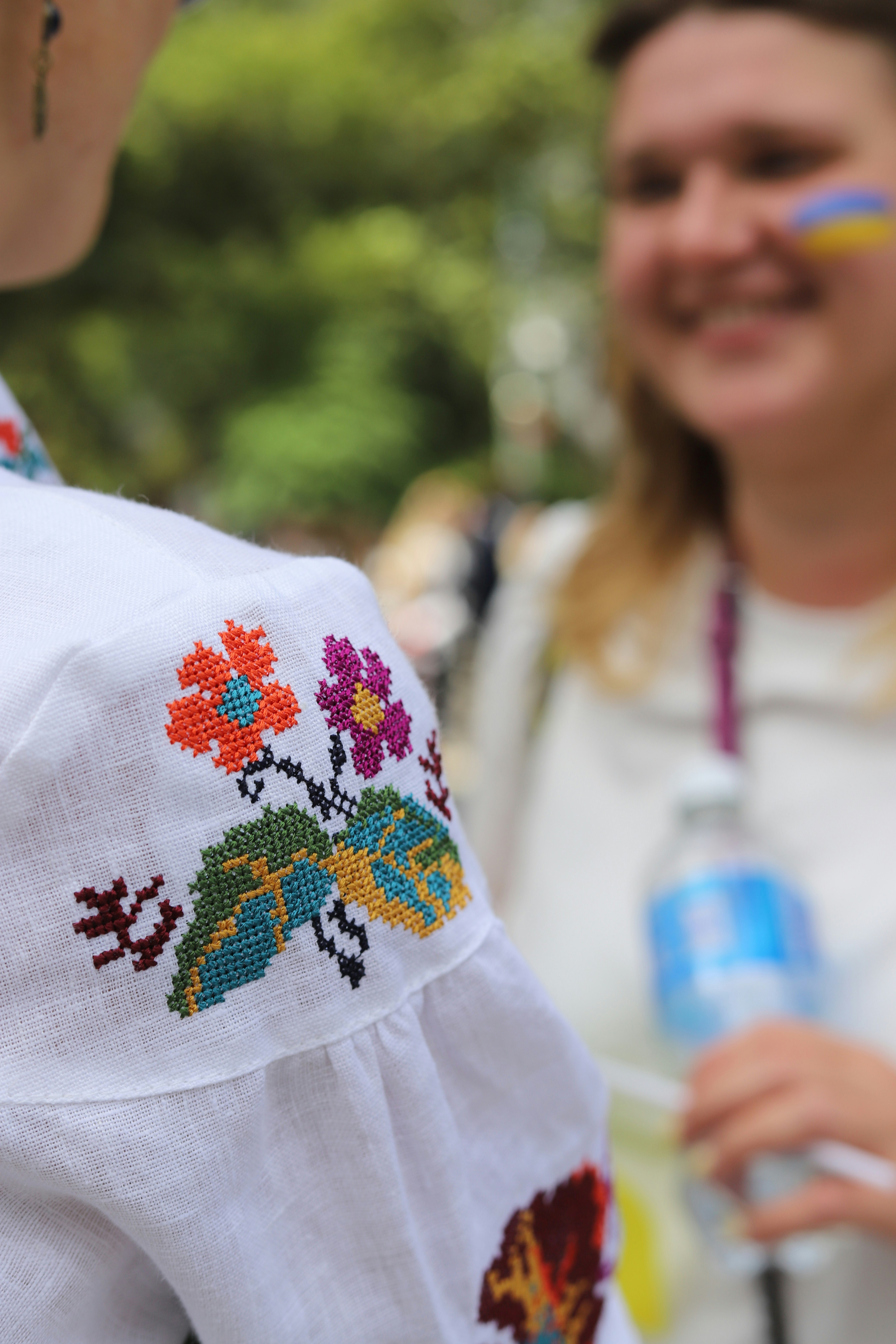 Close-up of colorful cross-stitch embroidery on a white linen sleeve, showing a teal and green butterfly beside orange and magenta flowers. In the soft blurred background, a smiling woman with blue and yellow face paint holds a water bottle. People gather in downtown Manhattan for a Ukrainian rally, waving blue and yellow flags and showing solidarity and support for Ukraine in the streets of New York City. Photo: Olek Buzunov (@buzunov.photoart)