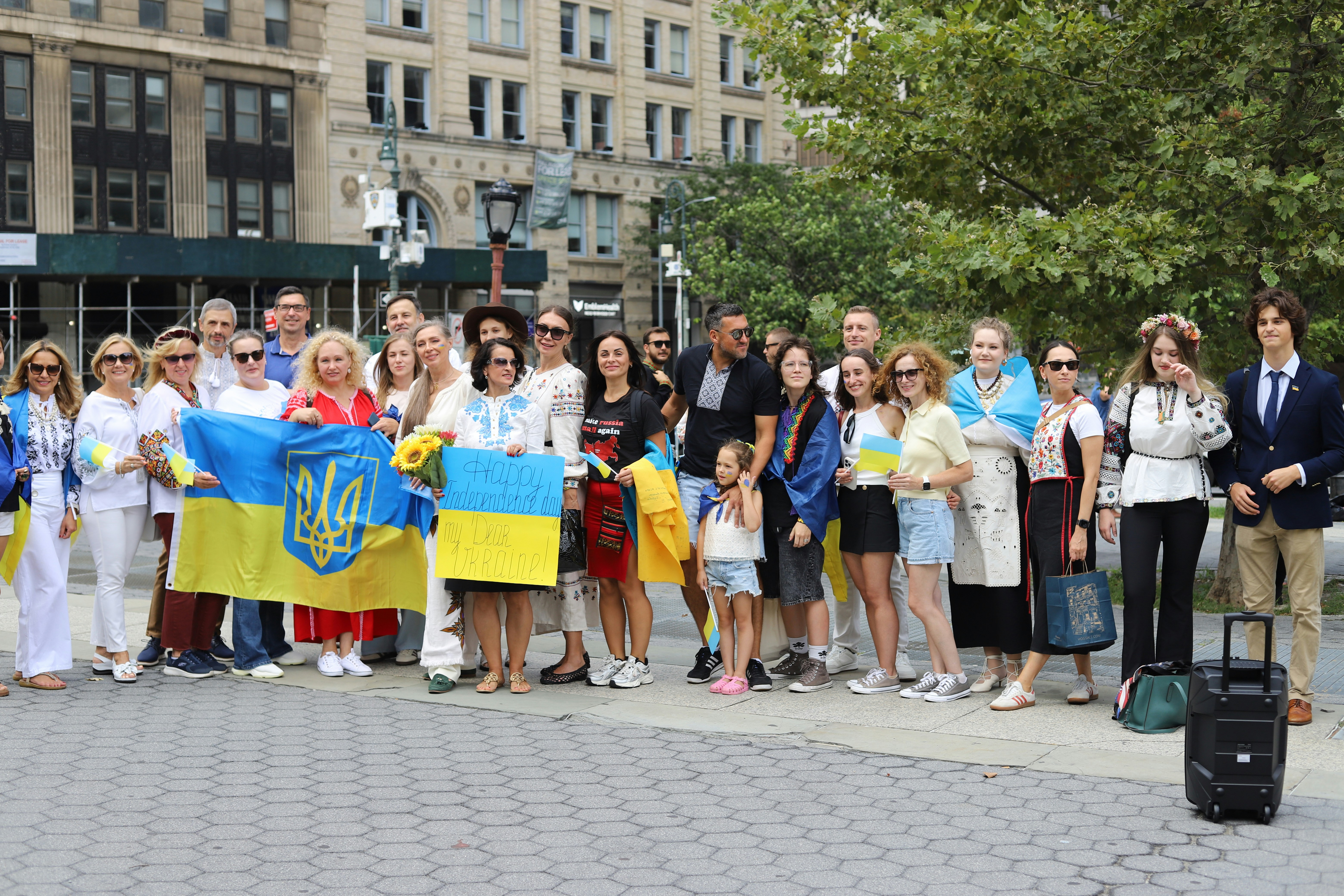 Rally participants stand shoulder to shoulder in downtown Manhattan beneath a leafy tree and a classic stone building. They wear traditional embroidered clothing, carry small Ukrainian flags and sunflowers, and a young man in a navy blazer and tie stands at the far right. People gather in downtown Manhattan for a Ukrainian rally, waving blue and yellow flags and showing solidarity and support for Ukraine in the streets of New York City. Photo: Olek Buzunov (@buzunov.photoart)