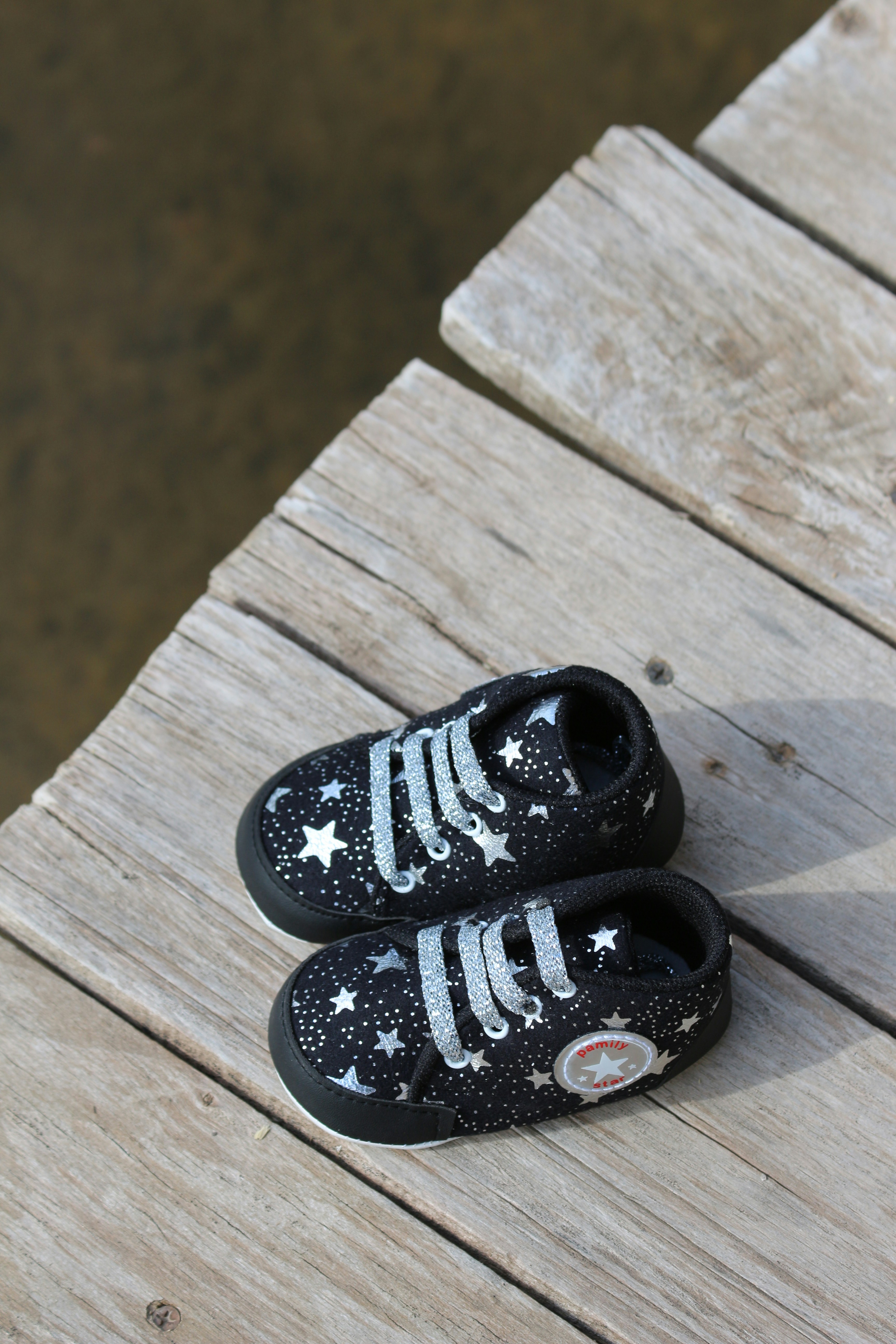 A pair of small black children's sneakers with silver star details resting on a sunlit wooden surface near the water.