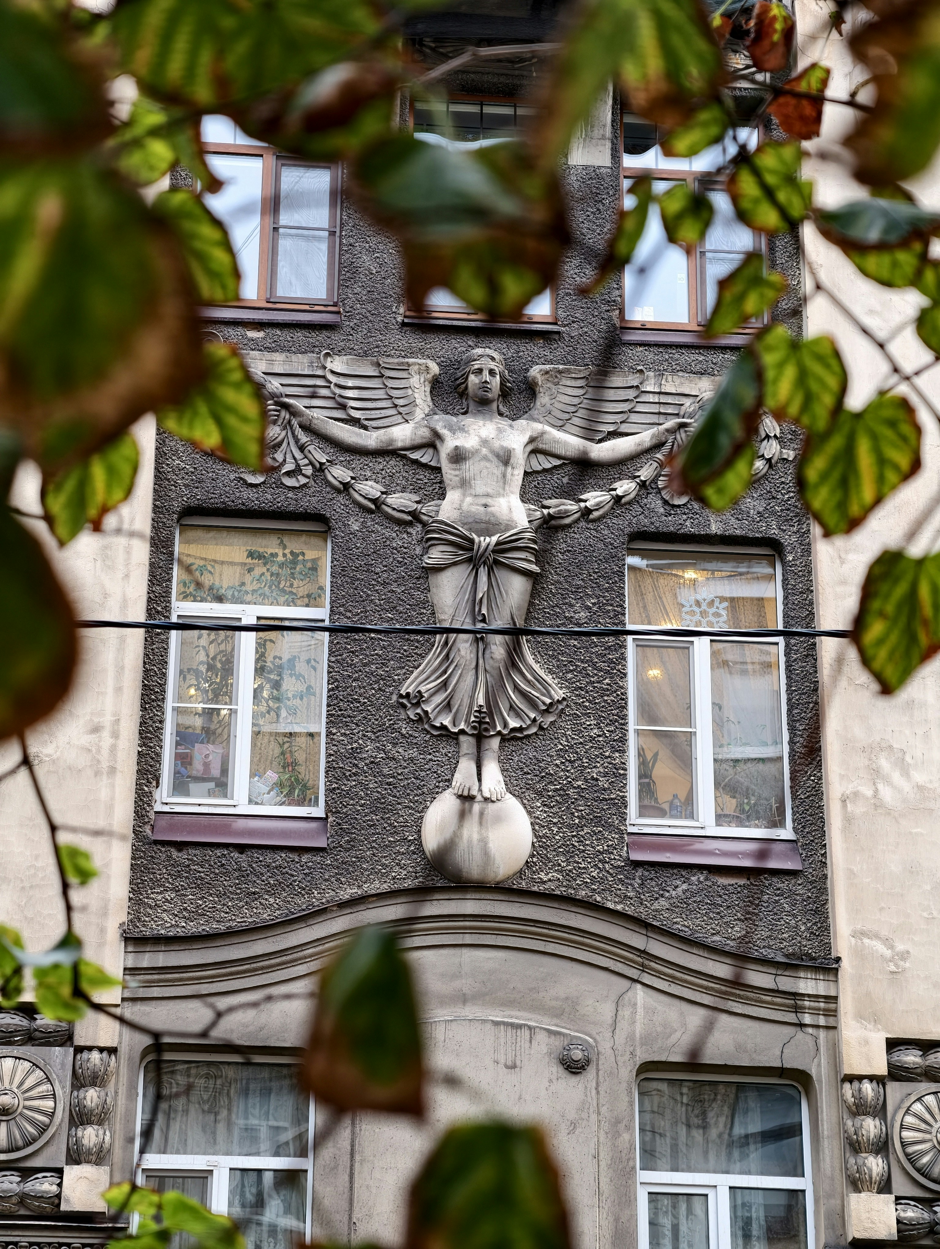 Decorative stone sculpture of a winged figure atop a textured building facade, framed by foreground leaves and windows below. Architectural detail and subtle human form captured through a street-level view.