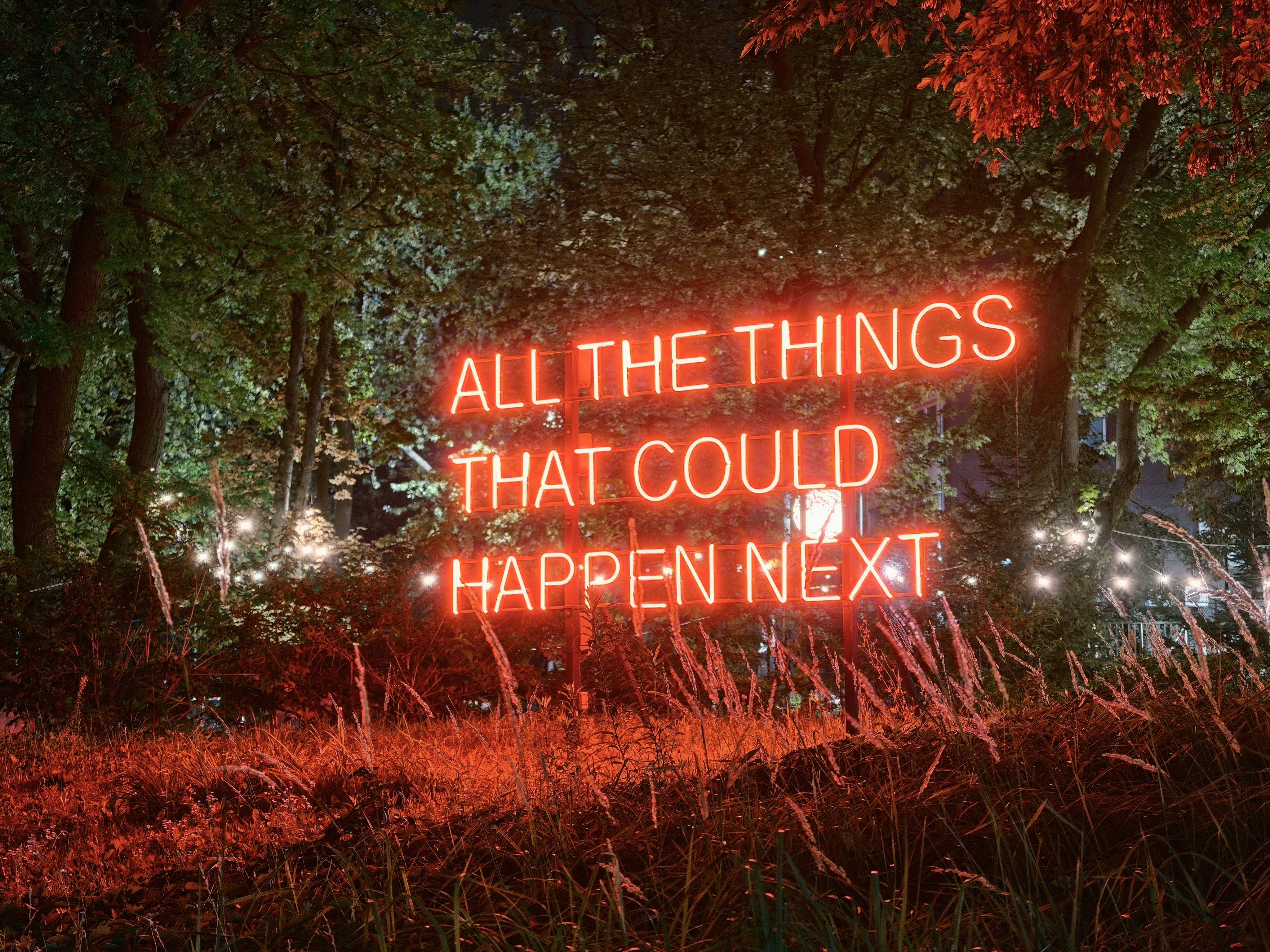 A red neon installation reading "ALL THE THINGS THAT COULD HAPPEN NEXT" by Tim Etchells glows among the trees at the corner of Emilii Plater and Nowogrodzka street in central Warsaw (Warszawa), Poland. Shot at night on an Oppo Find X9 Ultra with its OmniVision OV52A 1/1.28" sensor (70mm equivalent, F/7.6), the frame captures the neon's red light washing over tall grass, with the string lights of a nearby café terrace twinkling behind the foliage. Created in 2020 for Teatr Komuna Warszawa, the artwork turns an ordinary evening into a quiet meditation on the future.
