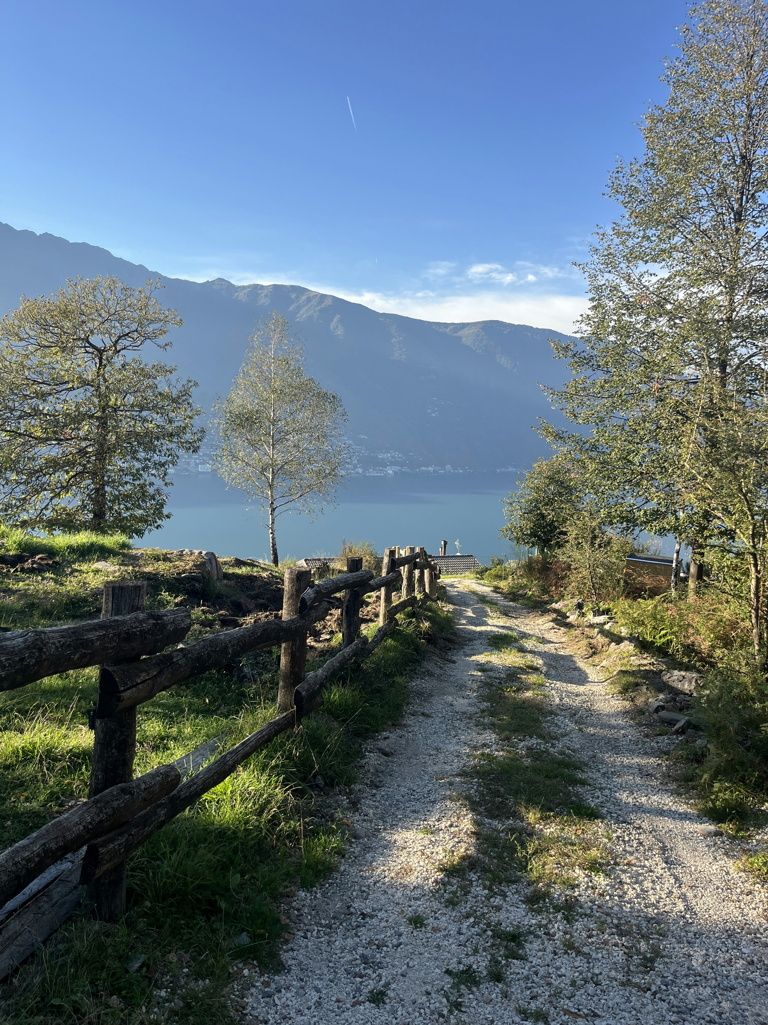 Gravel path with rustic wooden fence runs toward a tranquil lake and mountain range under a clear blue sky.