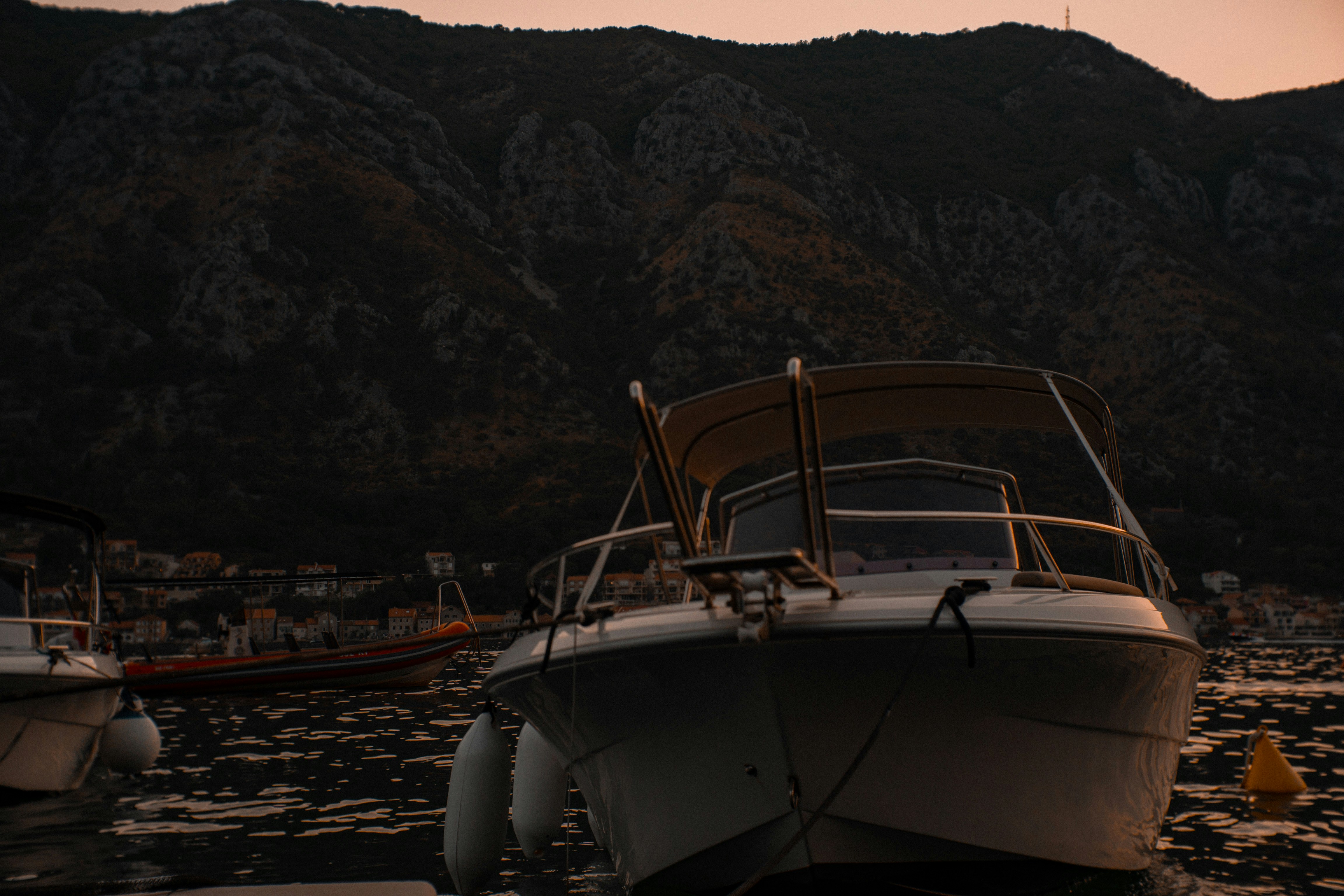Boats anchored in a calm harbor at dusk with a mountainous backdrop and mooring lines. A moored yacht dominates the foreground, creating a moody, low-light scene.