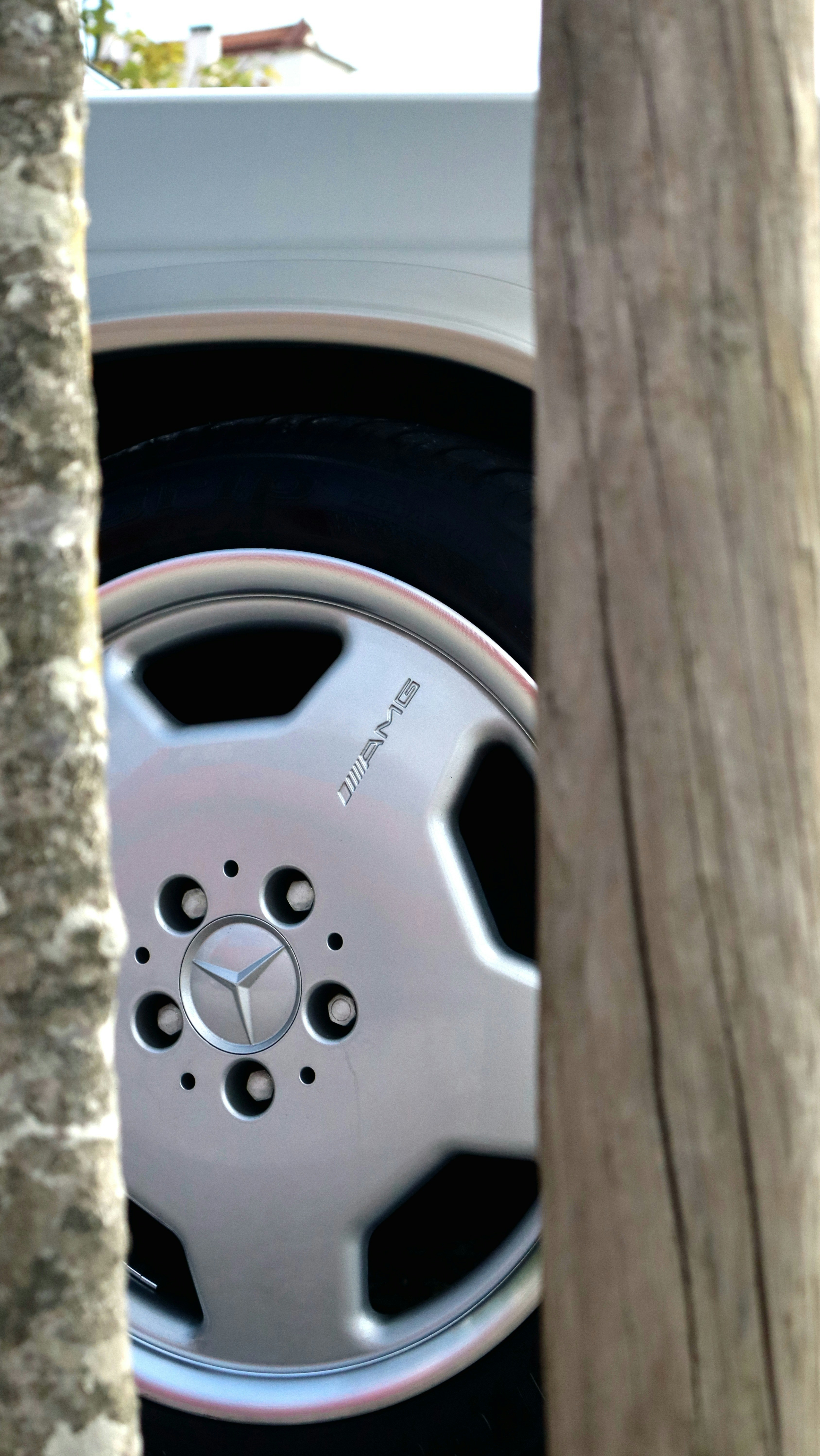 Silver Mercedes wheel partially visible between two wooden slats, suggesting a glimpse of a car behind a fence; a candid, architectural still focusing on texture and contrast.