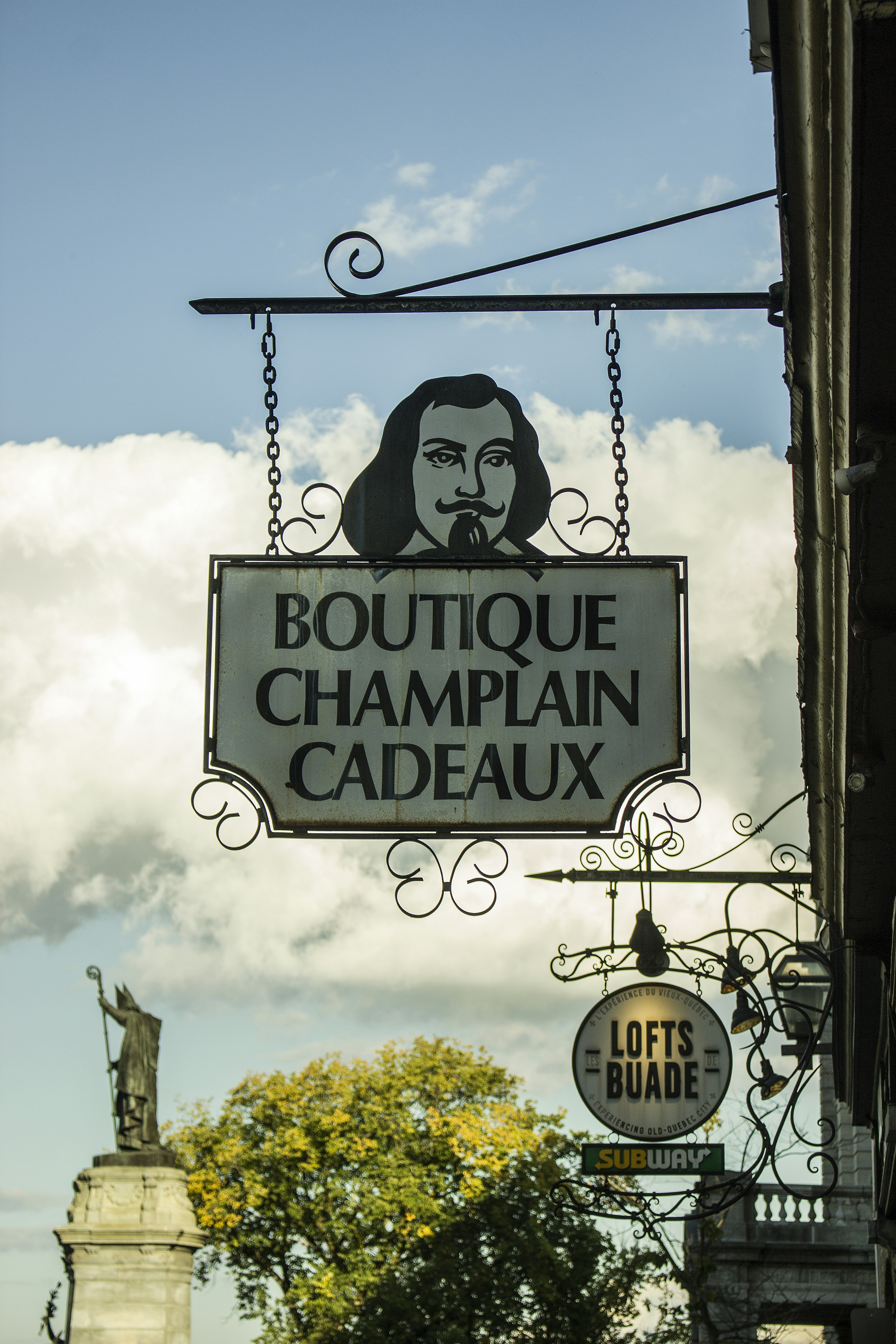 A decorative hanging sign reading 'Boutique Champlain Cadeaux' with a stylized face atop, set against a bright sky and a secondary circular 'Lofts Buade' sign in the background.
