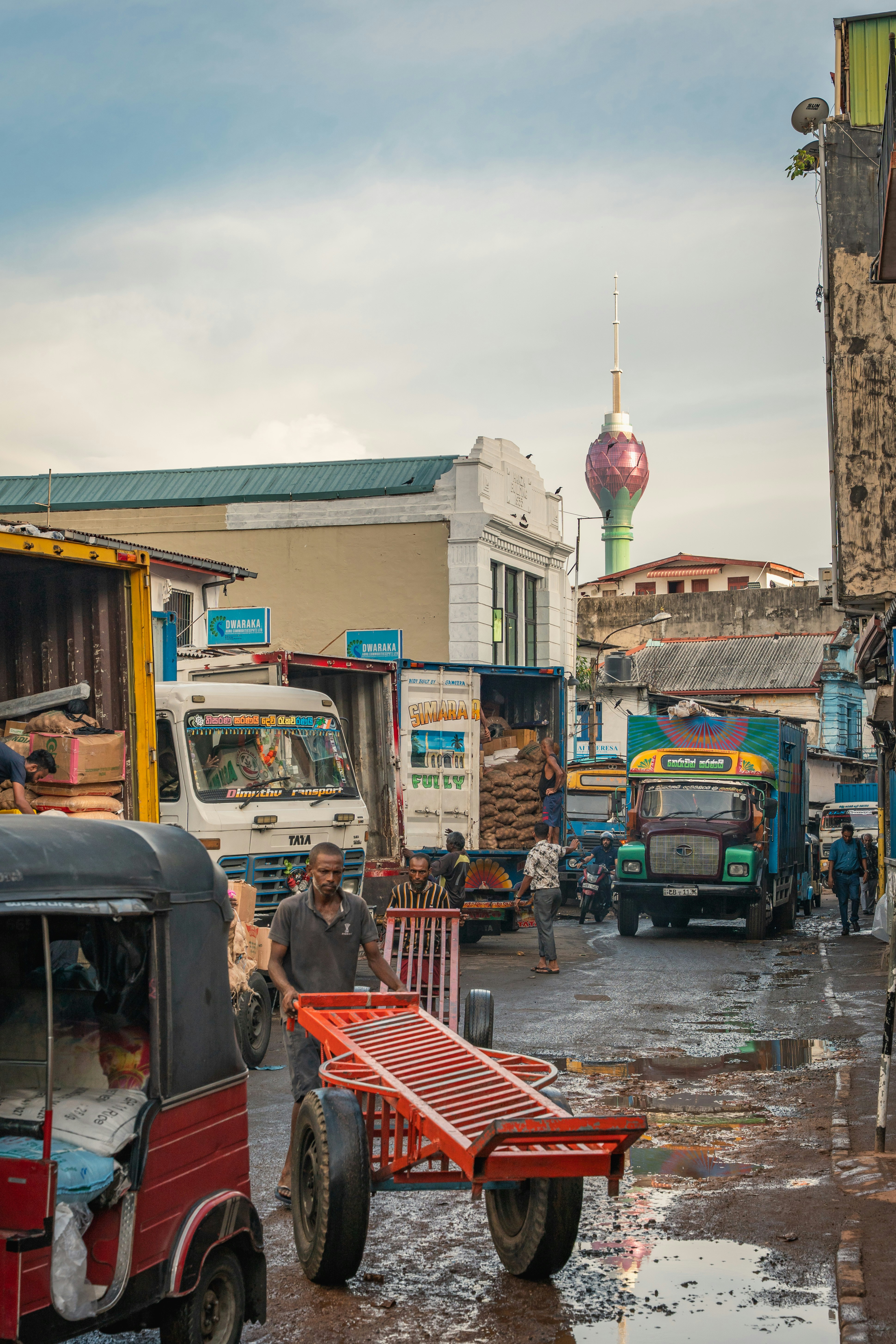 Market Lane in Motion: Rickshaws, Trucks, and Muddy Ground Under a Towered Skyline
