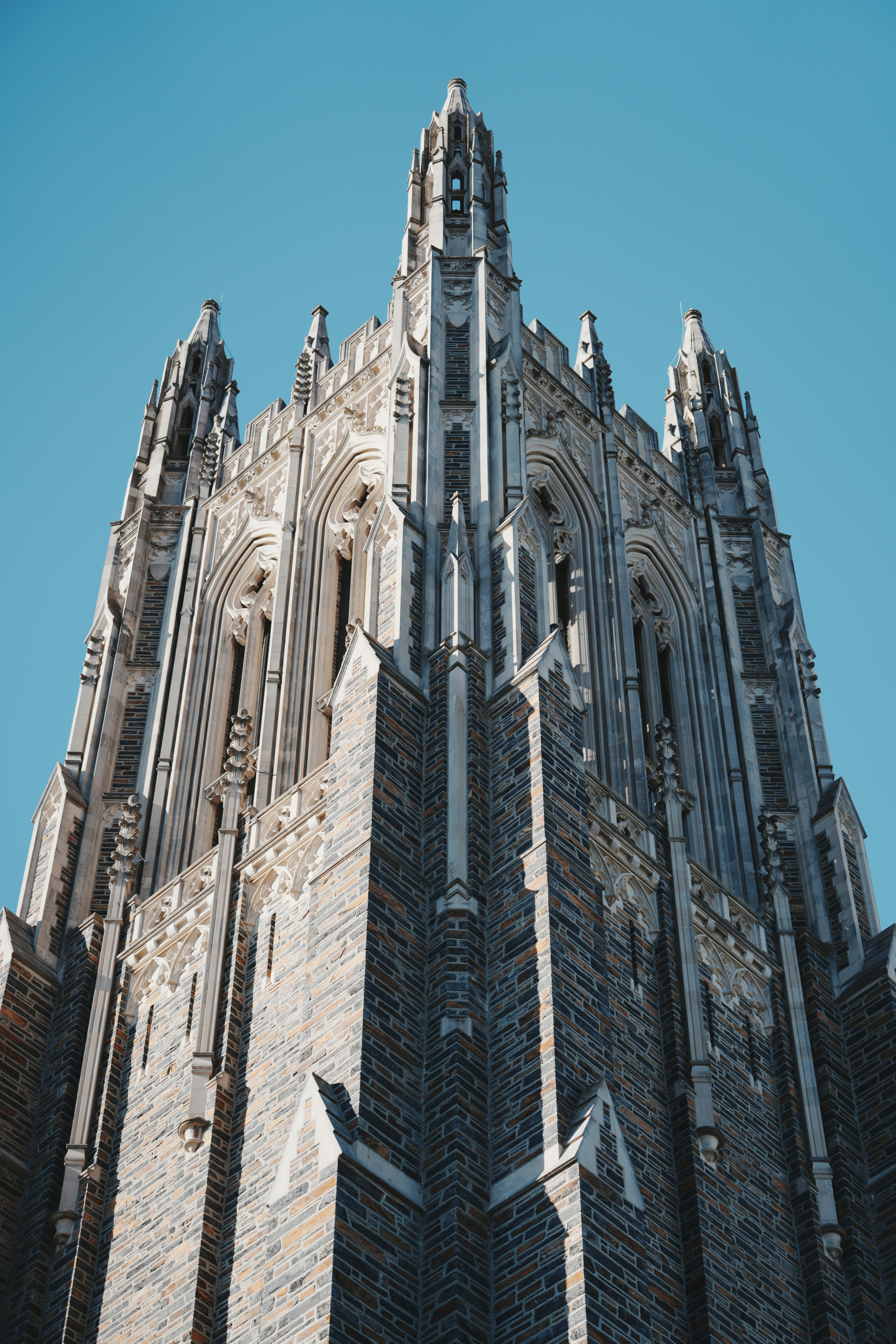 A towering Gothic stone spire rises against a bright blue sky, showcasing intricate buttresses and ornate stonework of a historic church façade.