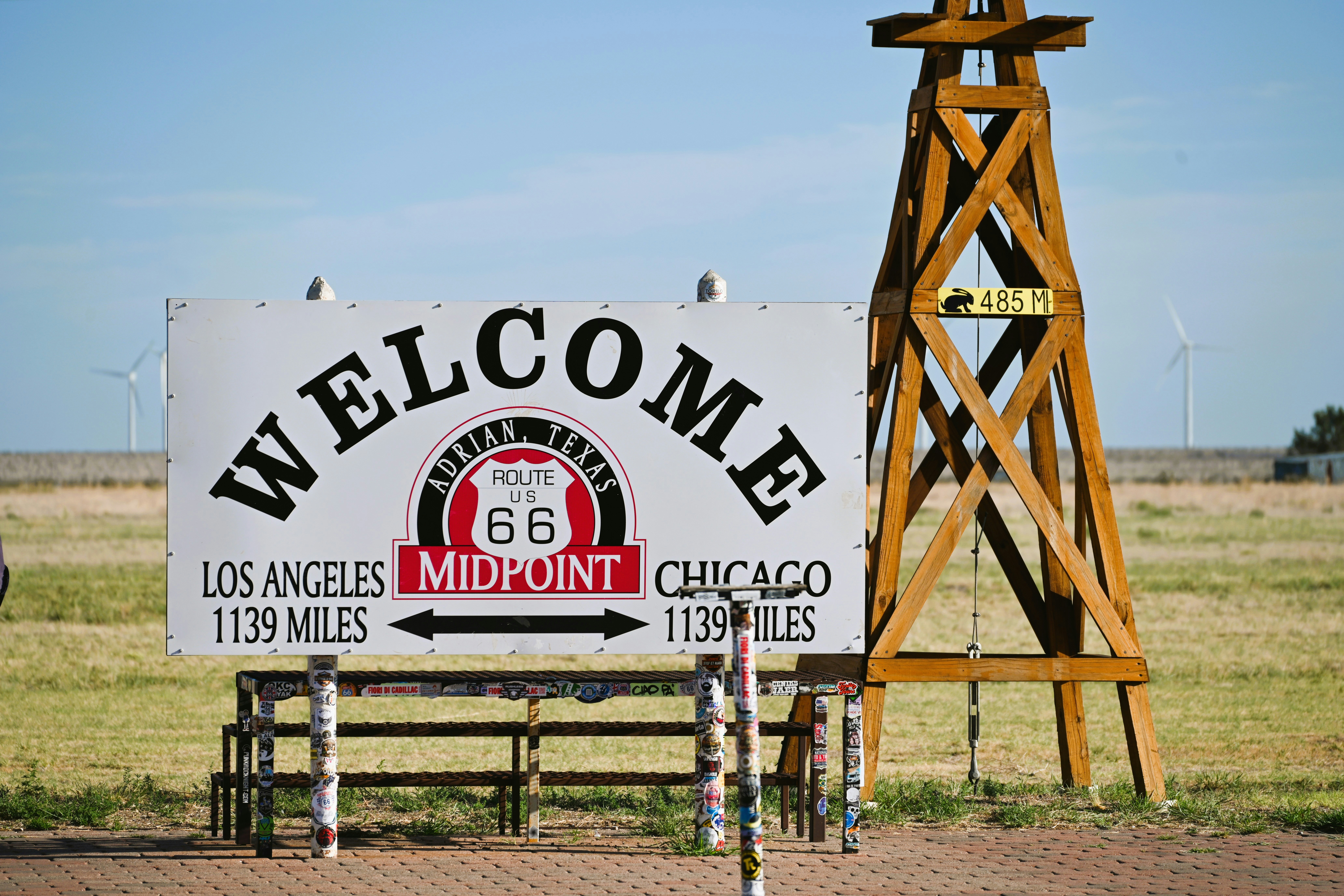 A large Welcome sign for Route 66 stands beside a tall wooden tower in an open prairie under a pale blue sky. The scene emphasizes travel nostalgia and the signage's weathered texture.