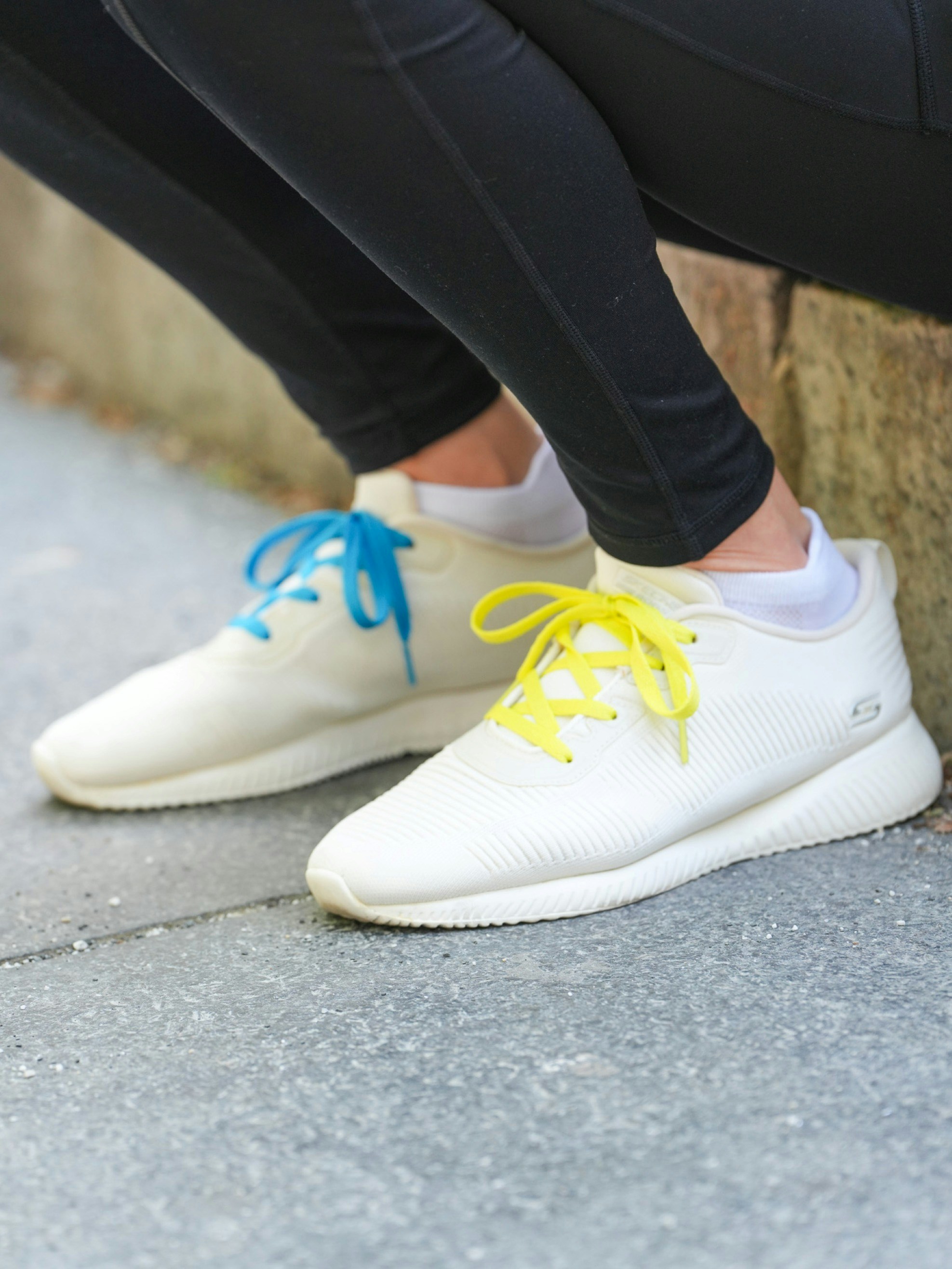 Close-up of white sneakers laced with one bright blue and one yellow shoelace, echoing the colors of the Ukrainian flag. The wearer, in black leggings and white socks, sits against a stone ledge above a gray pavement. Runners in traditional Ukrainian embroidered vyshyvanka shirts take part in a 2025 charity run in Central Park, celebrating Ukrainian culture and community while raising support for Ukraine. Photo: Olek Buzunov (@buzunov.photoart)
