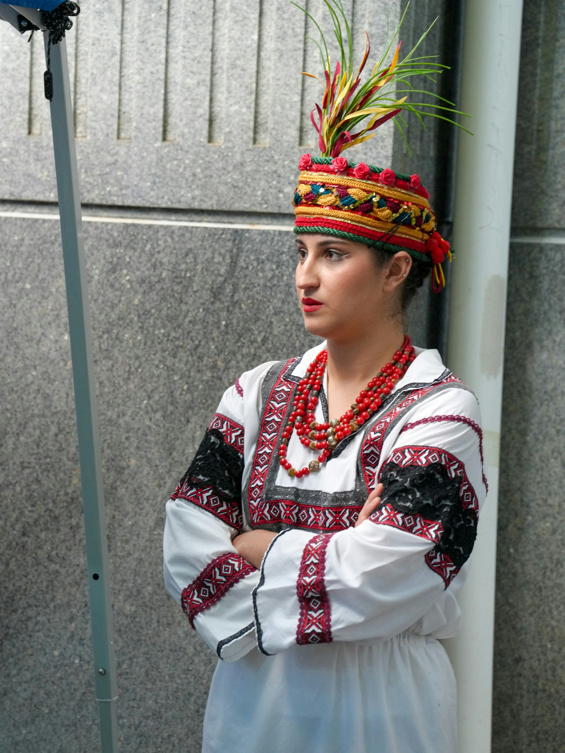 Colorful traditional headdress and embroidered white dress worn by a poised woman against a gray stone wall.