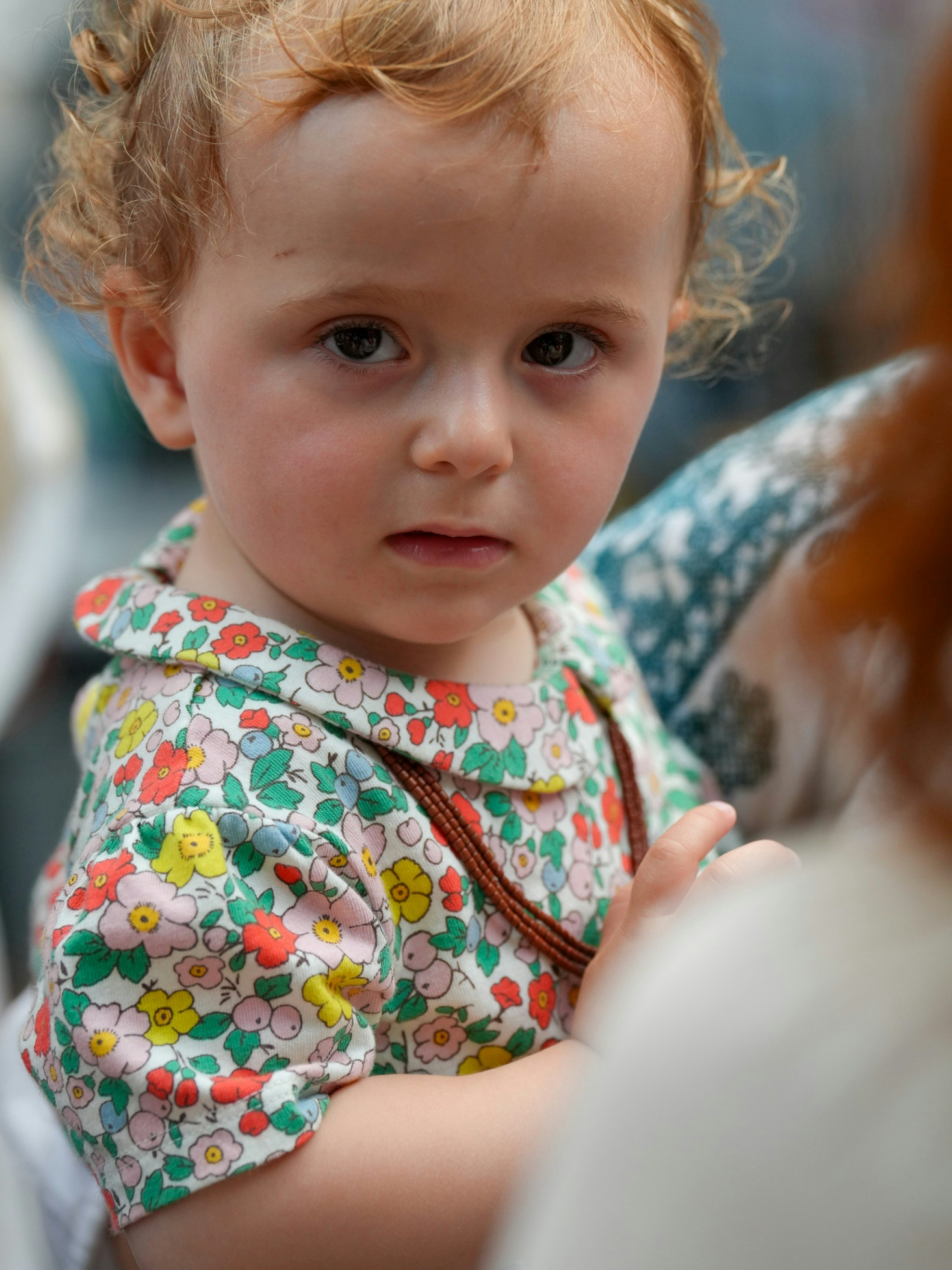 A curly red-haired toddler in a colorful floral print shirt with a Peter Pan collar looks seriously at the camera while being held in someone's arms. The crowd behind is softly blurred at the Ukrainian festival. Scenes from the 2025 Ukrainian Festival in Manhattan's East Village, celebrating Ukrainian culture, heritage, music, food, and community in the heart of Little Ukraine. Photo: Olek Buzunov (@buzunov.photoart)