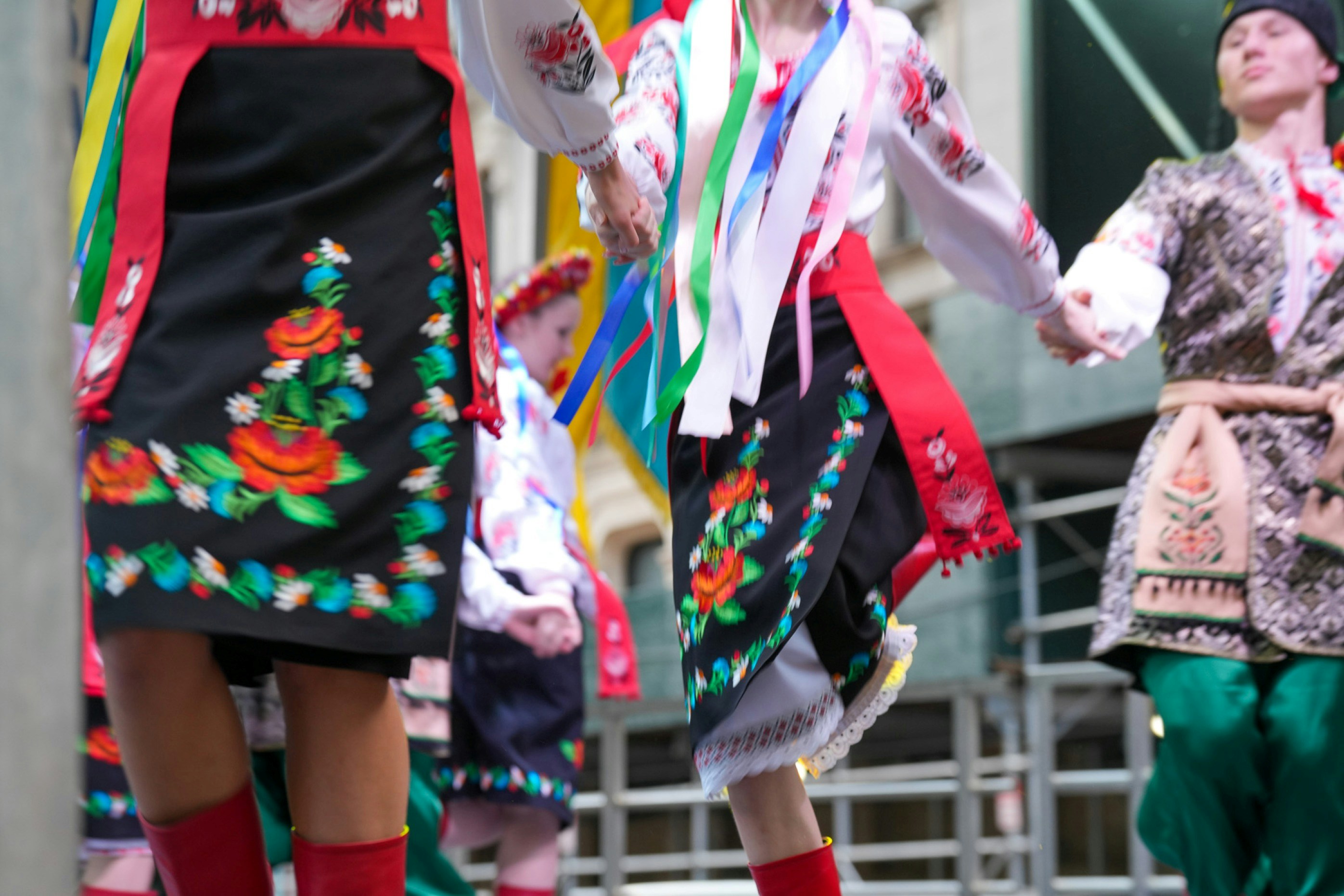 A low-angle view of folk dancers holding hands mid-step, their black skirts covered in bright embroidered flowers with red sashes and red boots. Colorful ribbons swirl as a young man in a brocade vest and green trousers dances at the edge of the stage. Scenes from the 2025 Ukrainian Festival in Manhattan's East Village, celebrating Ukrainian culture, heritage, music, food, and community in the heart of Little Ukraine. Photo: Olek Buzunov (@buzunov.photoart)