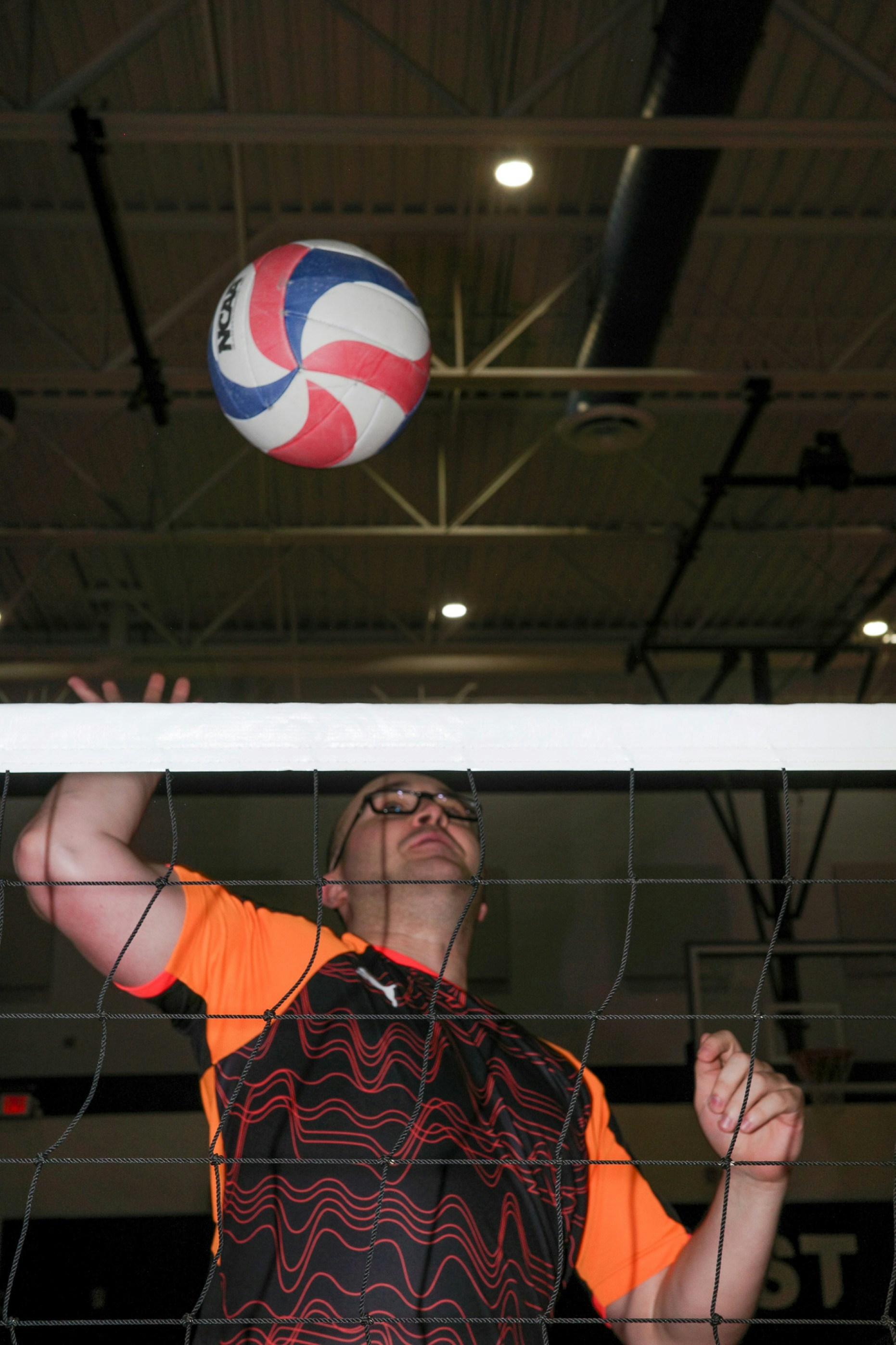 A man in glasses and an orange and black patterned jersey reaches up at the net as a red, white and blue volleyball flies overhead. The low angle shows the indoor gym's steel ceiling beams and lights above the white net tape. A friendly game of volleyball with a group of Ukrainian friends in Jersey City, New Jersey, celebrating community, friendship, and an active afternoon together outdoors. Photo: Olek Buzunov (@buzunov.photoart)