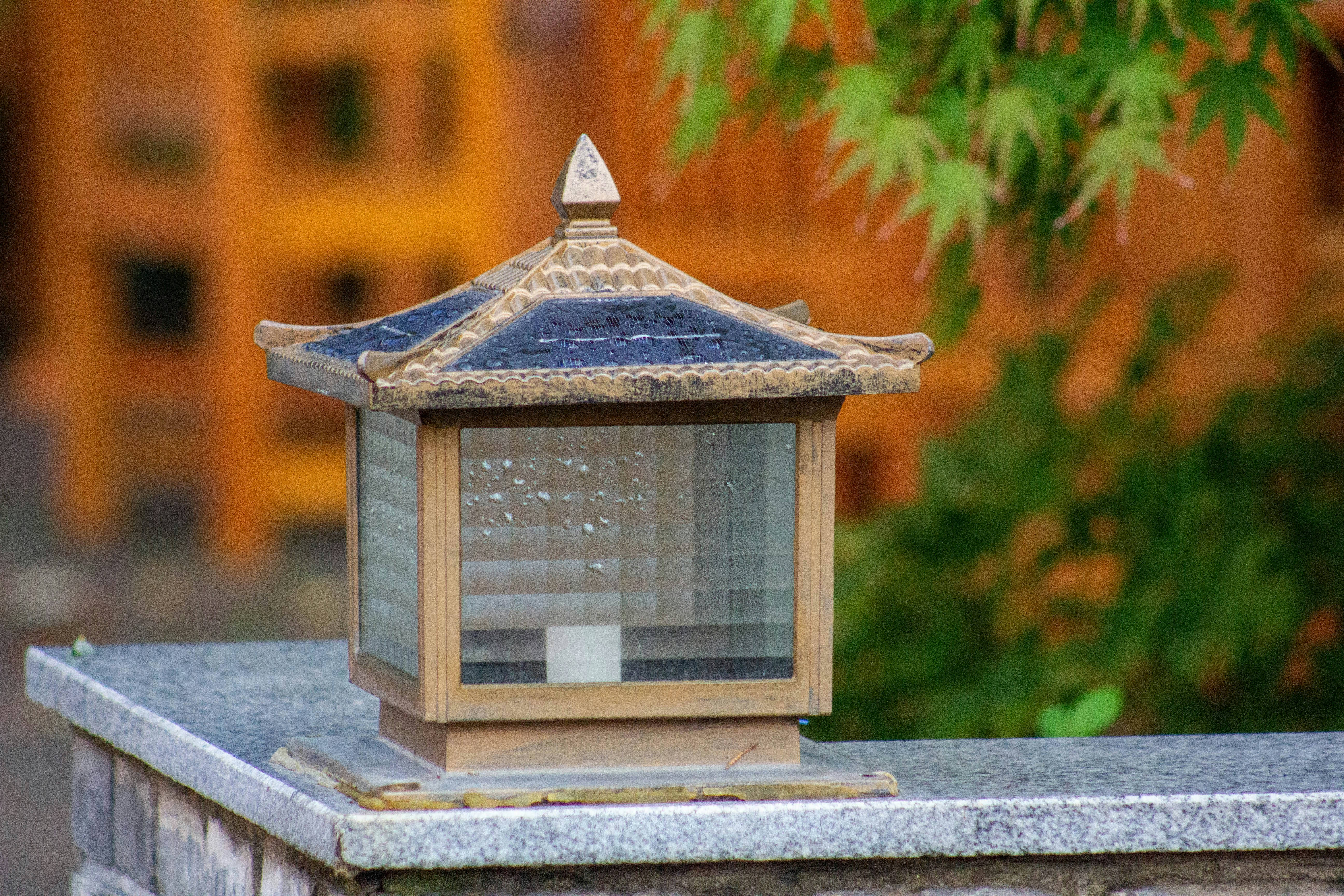 A small, traditional lantern sits on a stone ledge with blurred greenery and wooden architecture in the background, suggesting a tranquil courtyard scene.