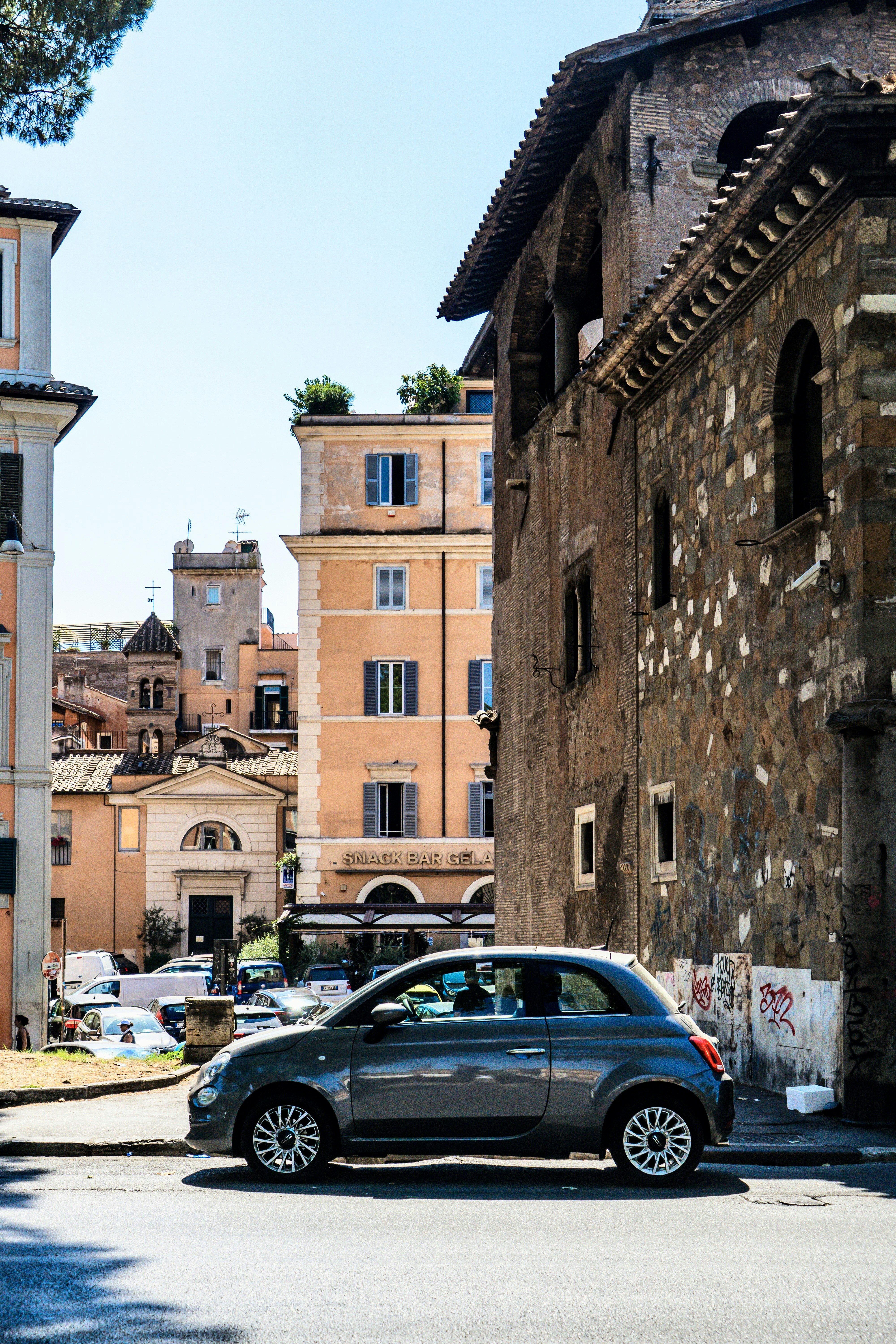 Blue hatchback in a sunlit Italian street framed by ancient stone and peach-colored facades