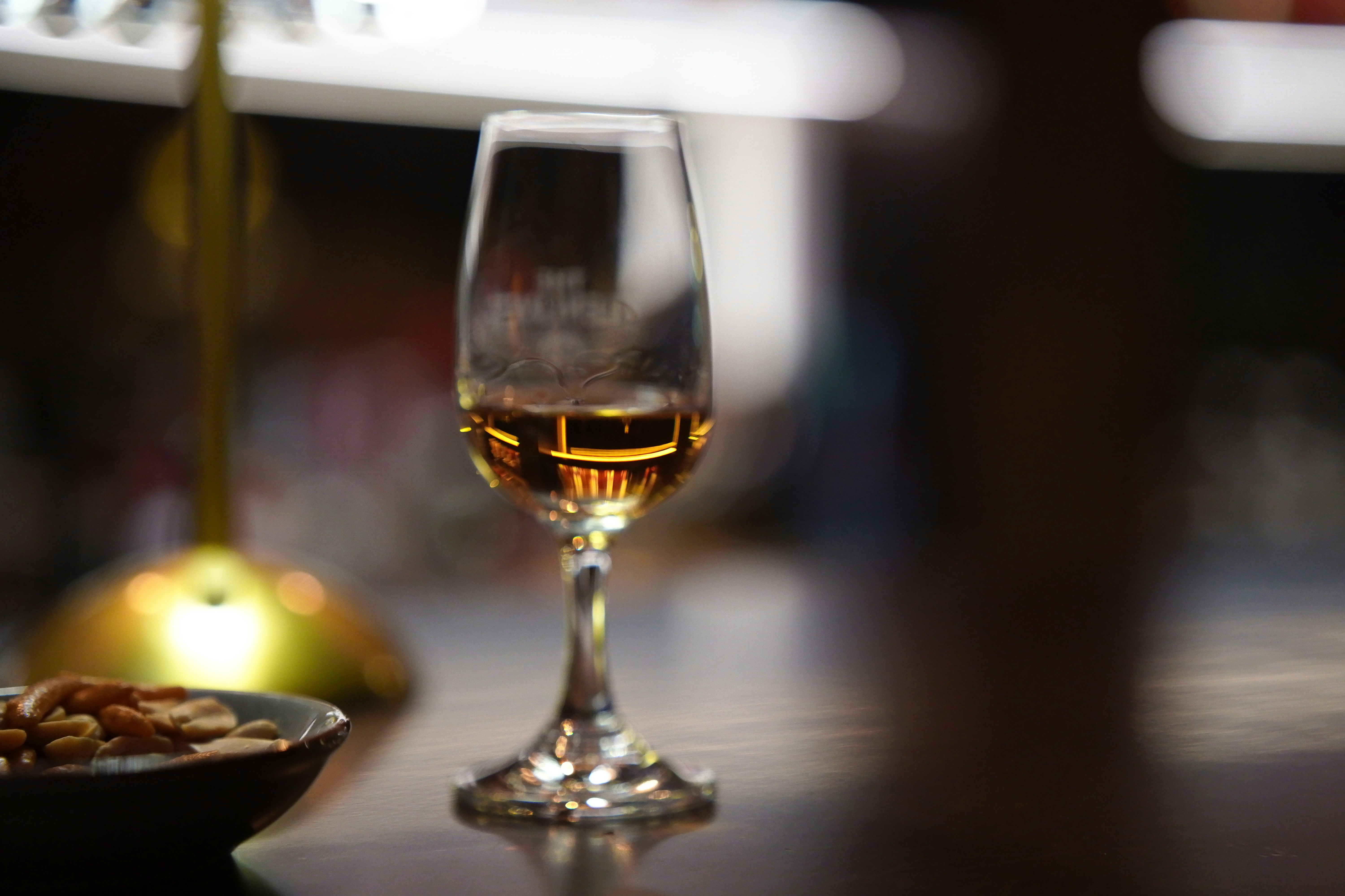 A single glass of amber beverage on a bar counter with soft bokeh and a nearby dish of nuts, evoking a quiet, intimate moment in a dim setting.