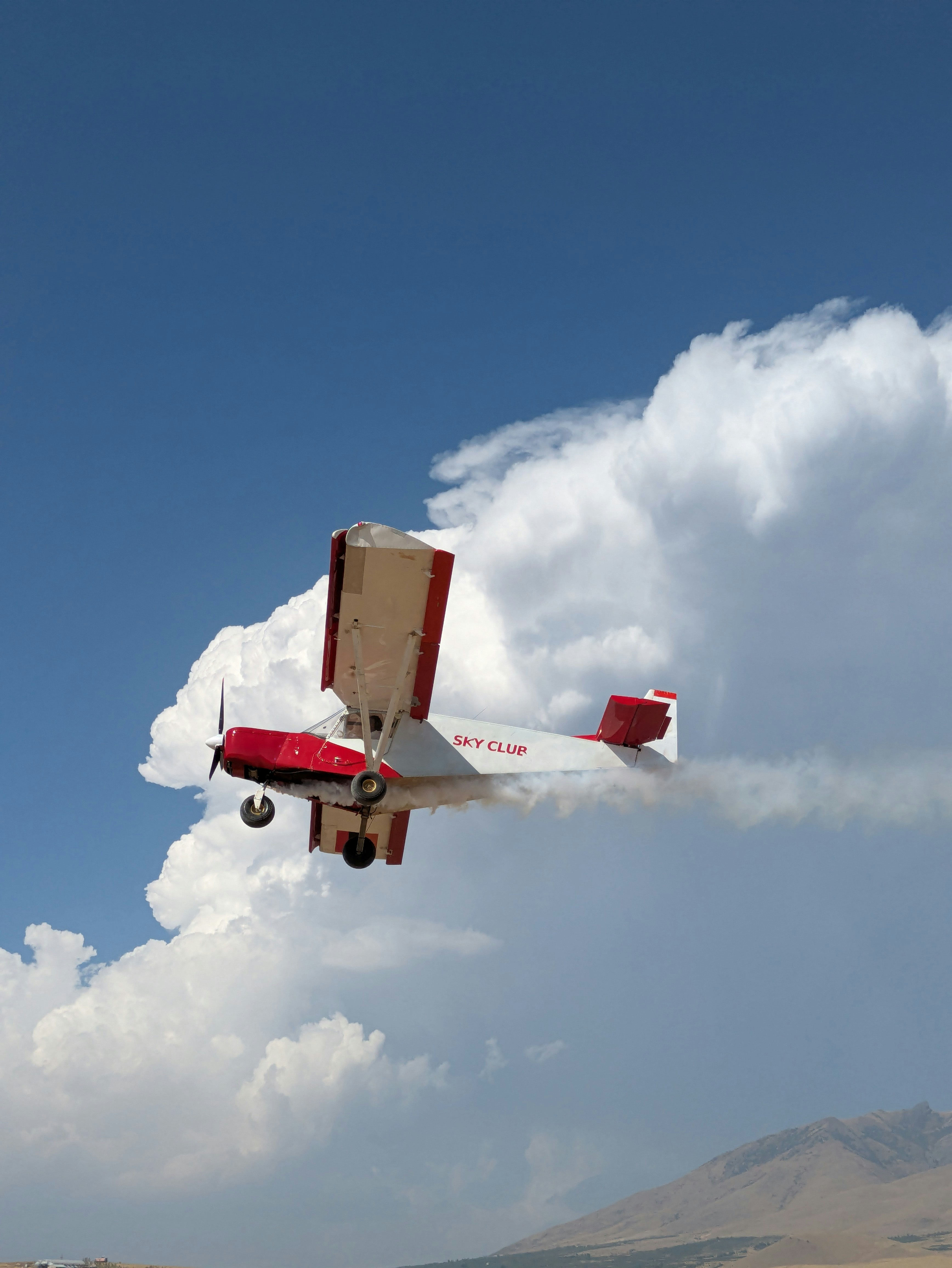 Small red-and-white plane climbs above terrain, leaving a white plume against a blue sky with distant hills.