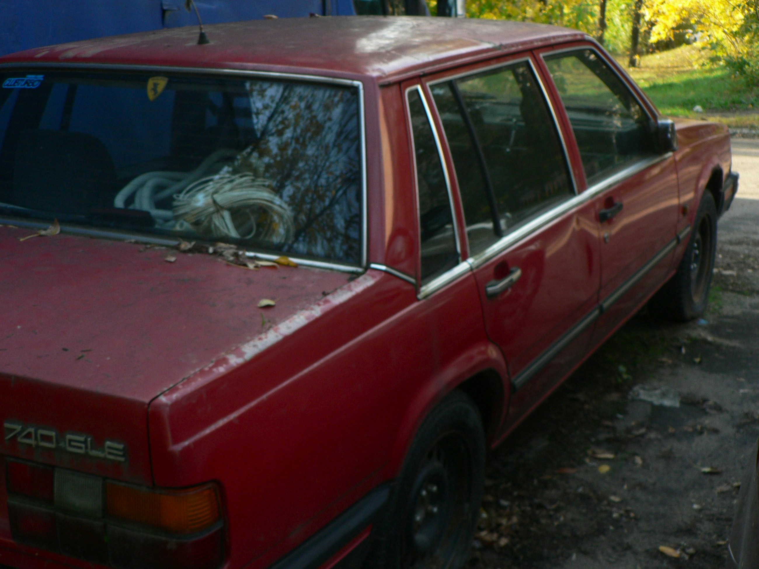 A rear three-quarter view of a weathered dark red Volvo 740 GLE sedan parked outdoors. The car's paint is faded and peeling, with fallen autumn leaves scattered across the trunk and rear window. Through the glass, a coil of white cable is visible inside, while the reflection of bare tree branches covers the rear windshield. Chrome trim runs along the windows and doors, contrasting with the black steel wheels and rubber bumper.