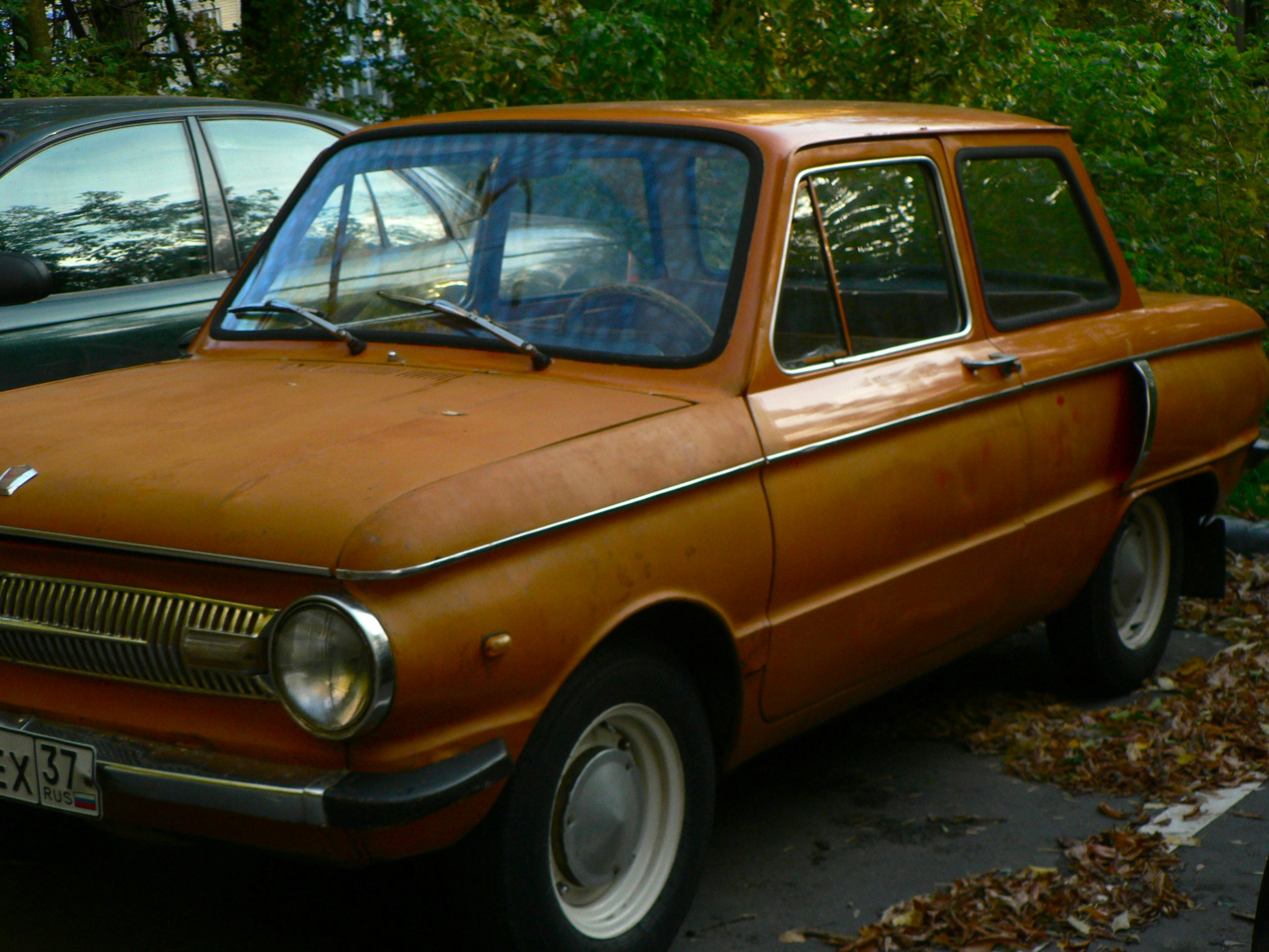 A close-up front view of a vintage orange sedan parked on a street covered with fallen autumn leaves. The car's retro design features a distinctive chrome grille, round headlights, and chrome trim running along the side. Reflections of trees are visible in the windshield, while a modern dark green car is parked partially visible behind it. The scene captures a nostalgic contrast between classic and contemporary automobiles on a quiet autumn day.