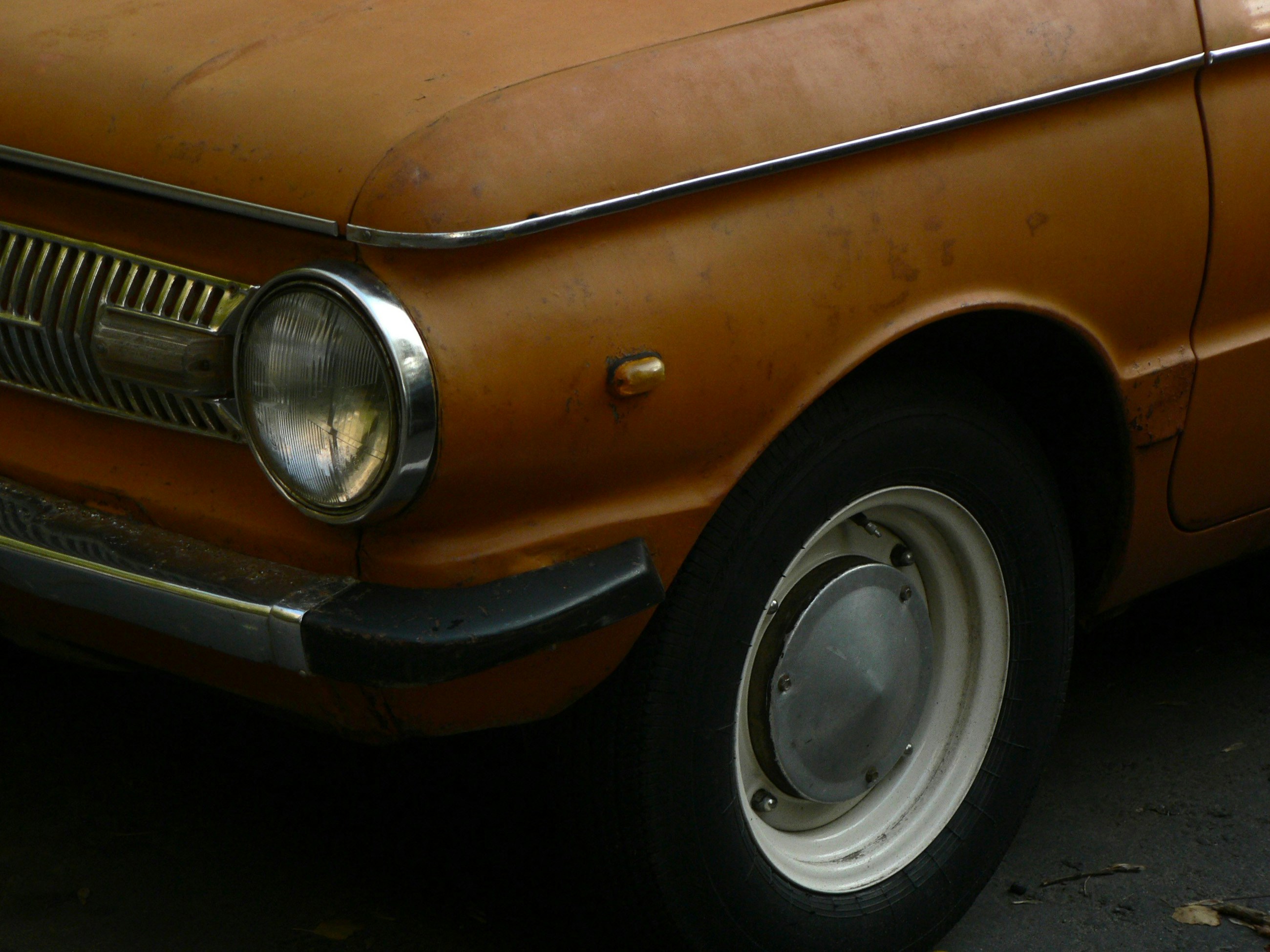 A detailed close-up of the front end of a vintage car, focusing on the round headlight with a chrome bezel, the ribbed chrome grille, and the black rubber bumper. The car's body is painted in a faded, weathered orange with patches of rust and dirt, showing its age. A simple steel wheel with a plain grey hubcap sits below a curved fender, while a small amber side marker light and a thin chrome trim line add classic details. The dark asphalt ground and muted lighting give the scene a quiet, nostalgic feel.
