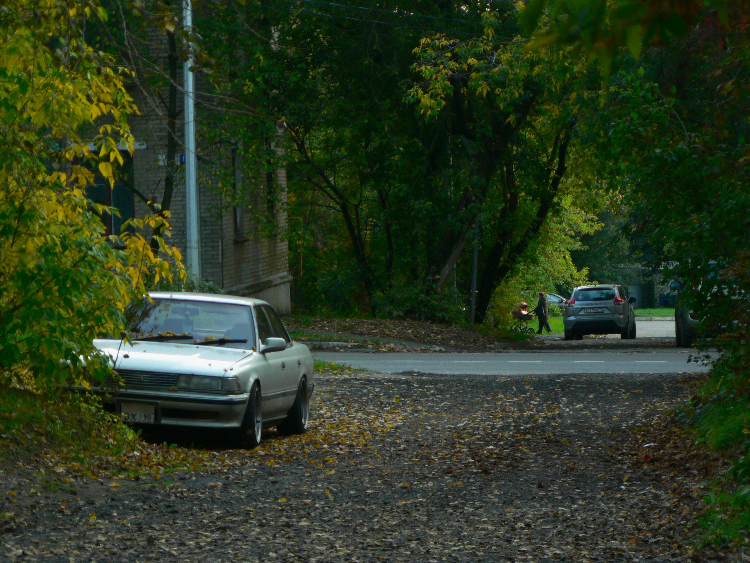A silver sedan parked on a leaf-strewn dirt path, framed by a large tree with bright yellow autumn foliage on the left and a brick building behind it. In the distance, a paved road crosses the scene, where a person walks with a baby stroller and a grey SUV is parked. Another car is partially visible on the far right, while dense green and yellow trees canopy the street, creating a quiet, everyday autumn atmosphere.
