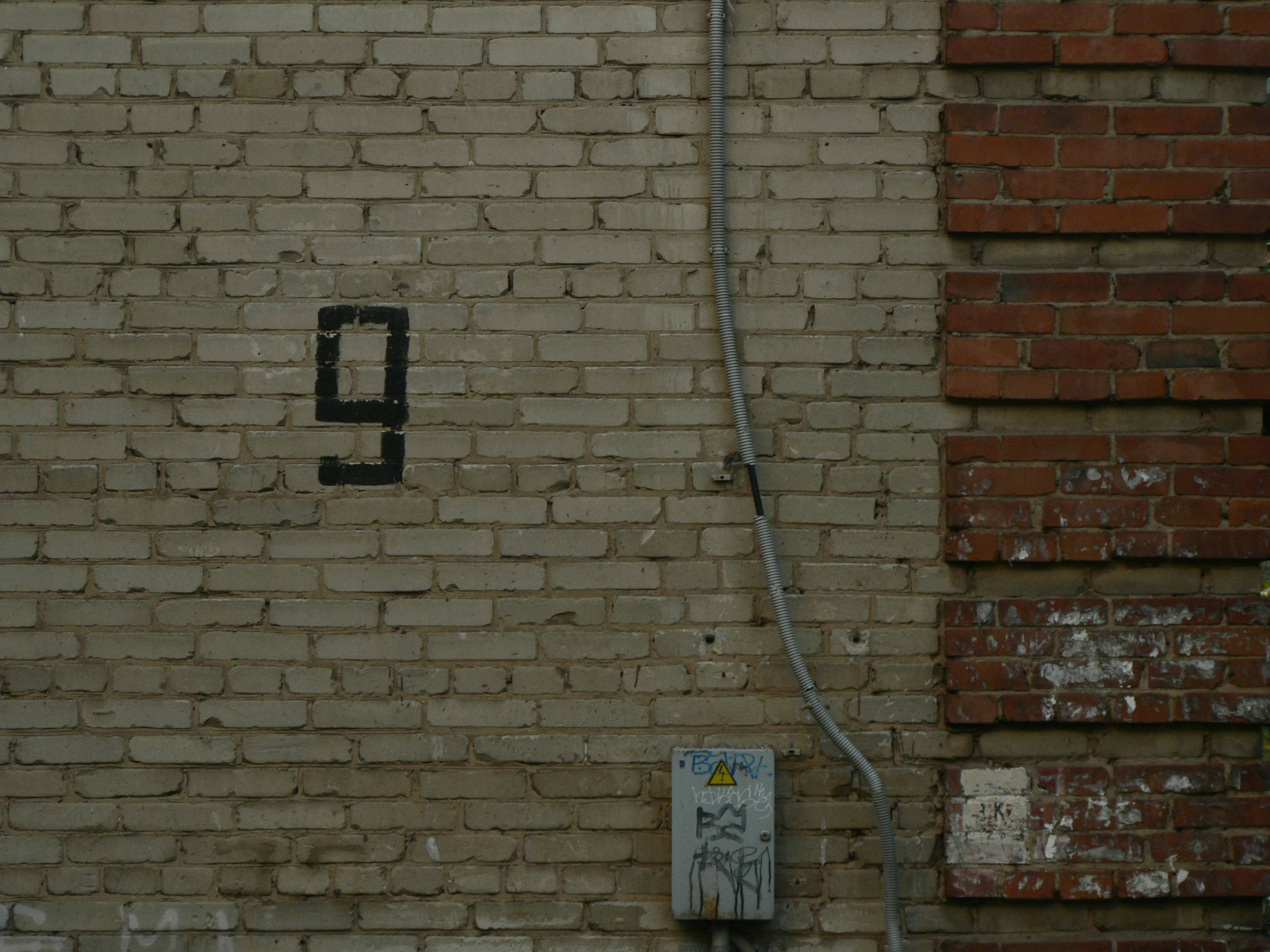 A textured section of an old brick wall, featuring a mix of light beige and reddish-brown bricks. On the left, a large, thick black number '9' is painted directly onto the masonry. A grey corrugated electrical conduit runs vertically down the center, connecting to a metal utility box at the bottom. The box is covered in graffiti, including a yellow warning triangle and black tags. The varied brickwork and utilitarian details create a gritty, industrial urban aesthetic.
