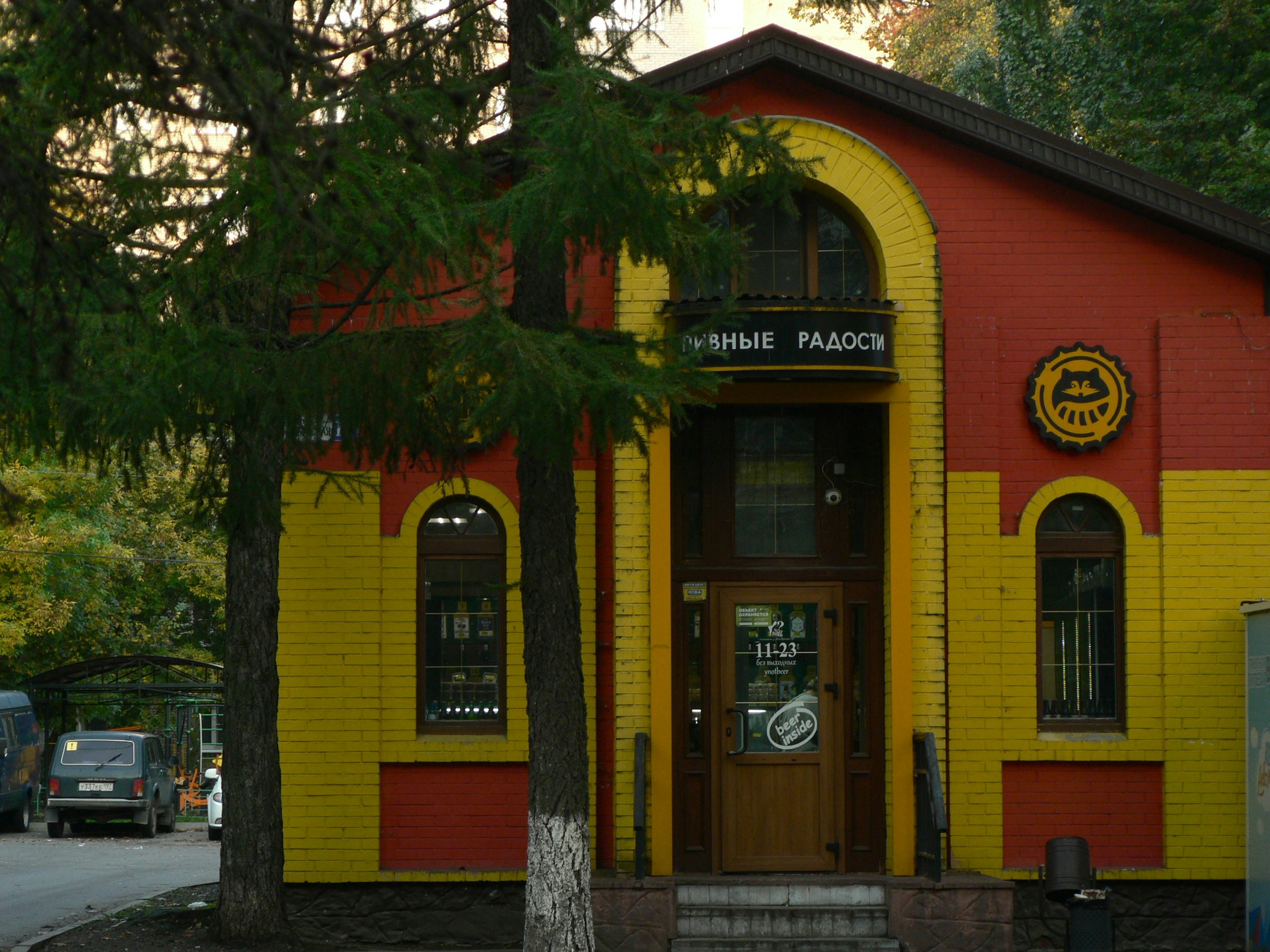 A brightly painted building, featuring a striking color-blocked facade of red and yellow brick. The central entrance is framed by a yellow arch and topped with a black sign reading 'ПИВНЫЕ РАДОСТИ' (Beer Joys) in white Cyrillic letters. To the right, a large circular logo displays a stylized black cat face on a yellow gear-like background. Arched windows flank the wooden double doors, which have a 'beer inside' sticker and display the opening hours '11-23'.