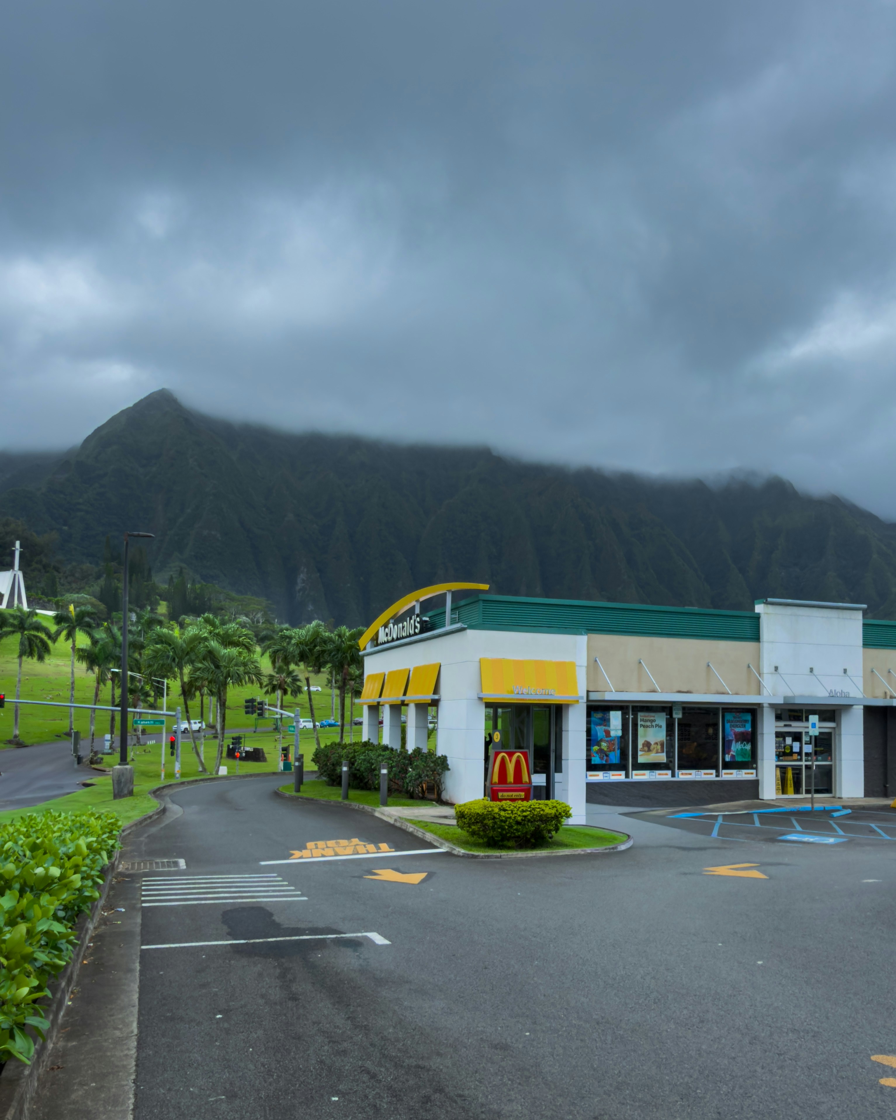 McDonald’s restaurant sits in a parking lot with a winding road and tropical landscaping, backed by a dramatic mountain range shrouded in clouds.